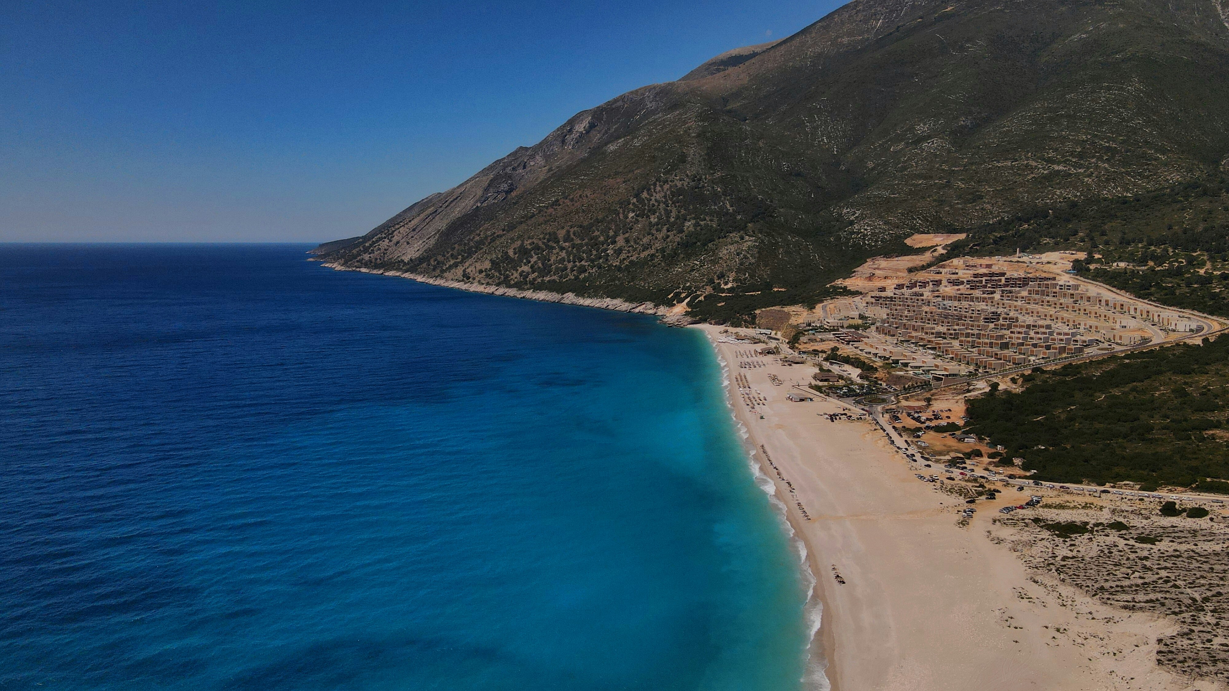 Aerial view of a pristine beach meeting the azure waters, bordered by a rugged mountain landscape and dotted with beachgoers.