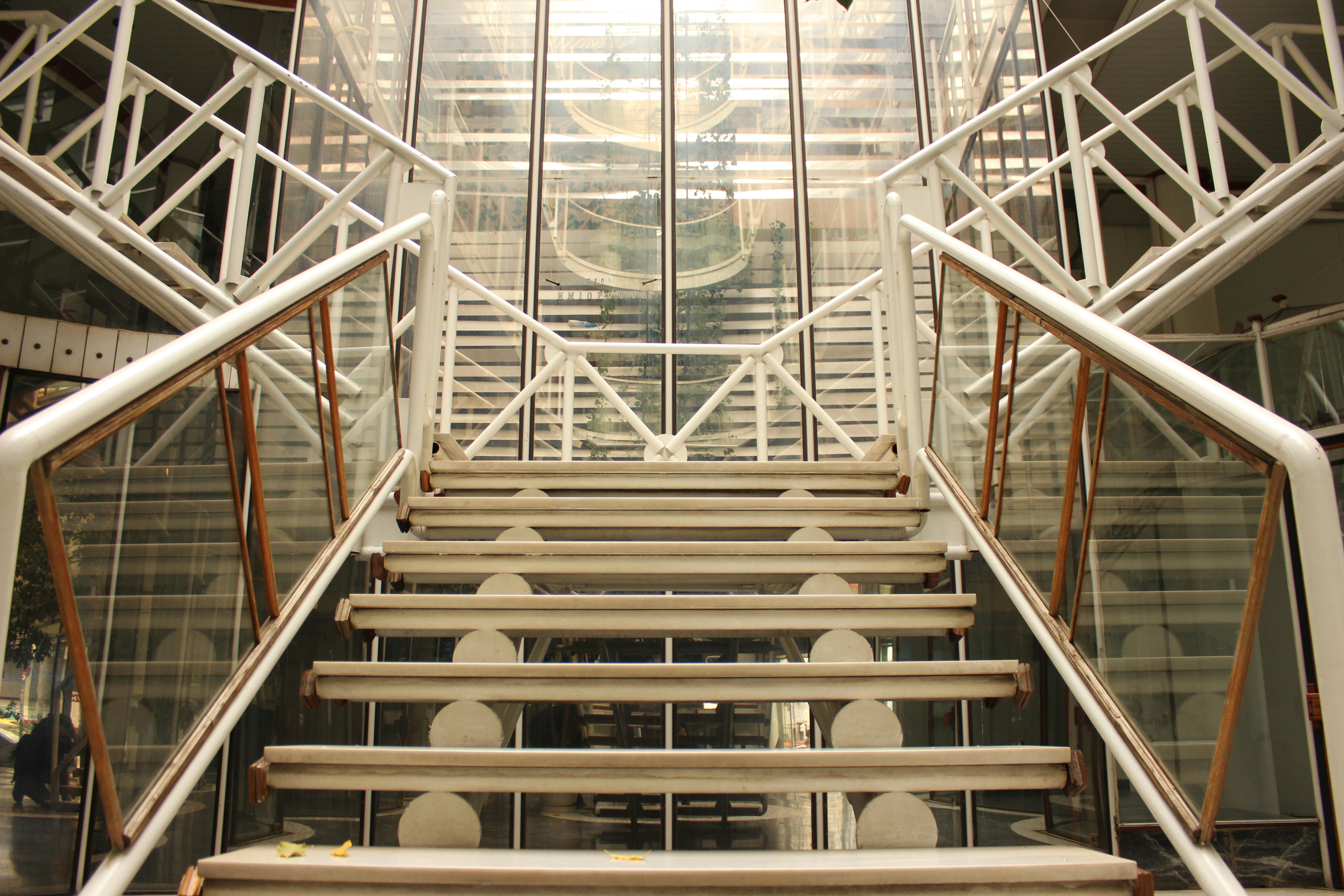 A modern staircase with white railings and circular steps leads upward, framed by glass walls that reflect light. The architectural design emphasizes depth and perspective.