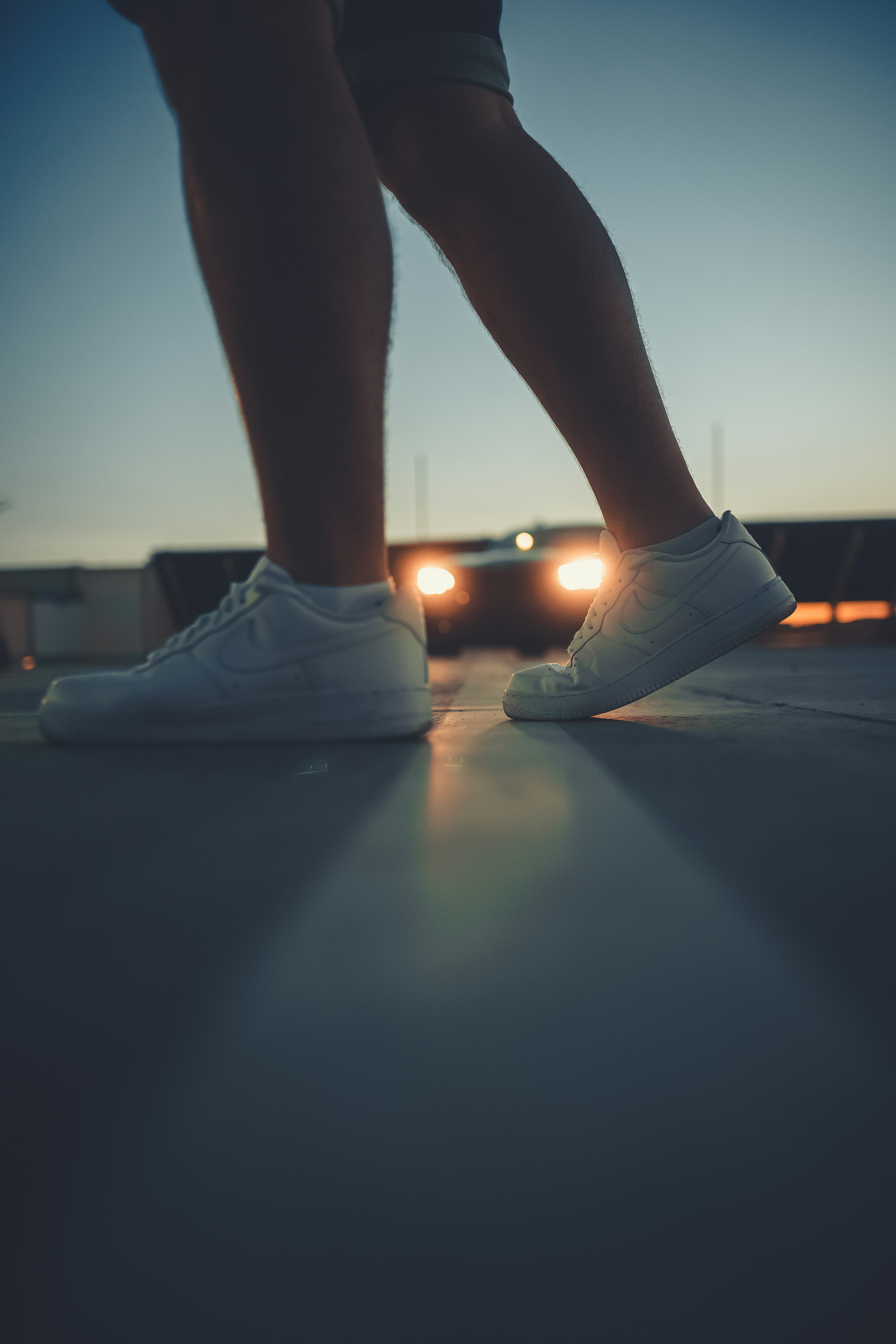 A person's legs in white sneakers stride across a parking lot at dusk, with headlights illuminating the background.