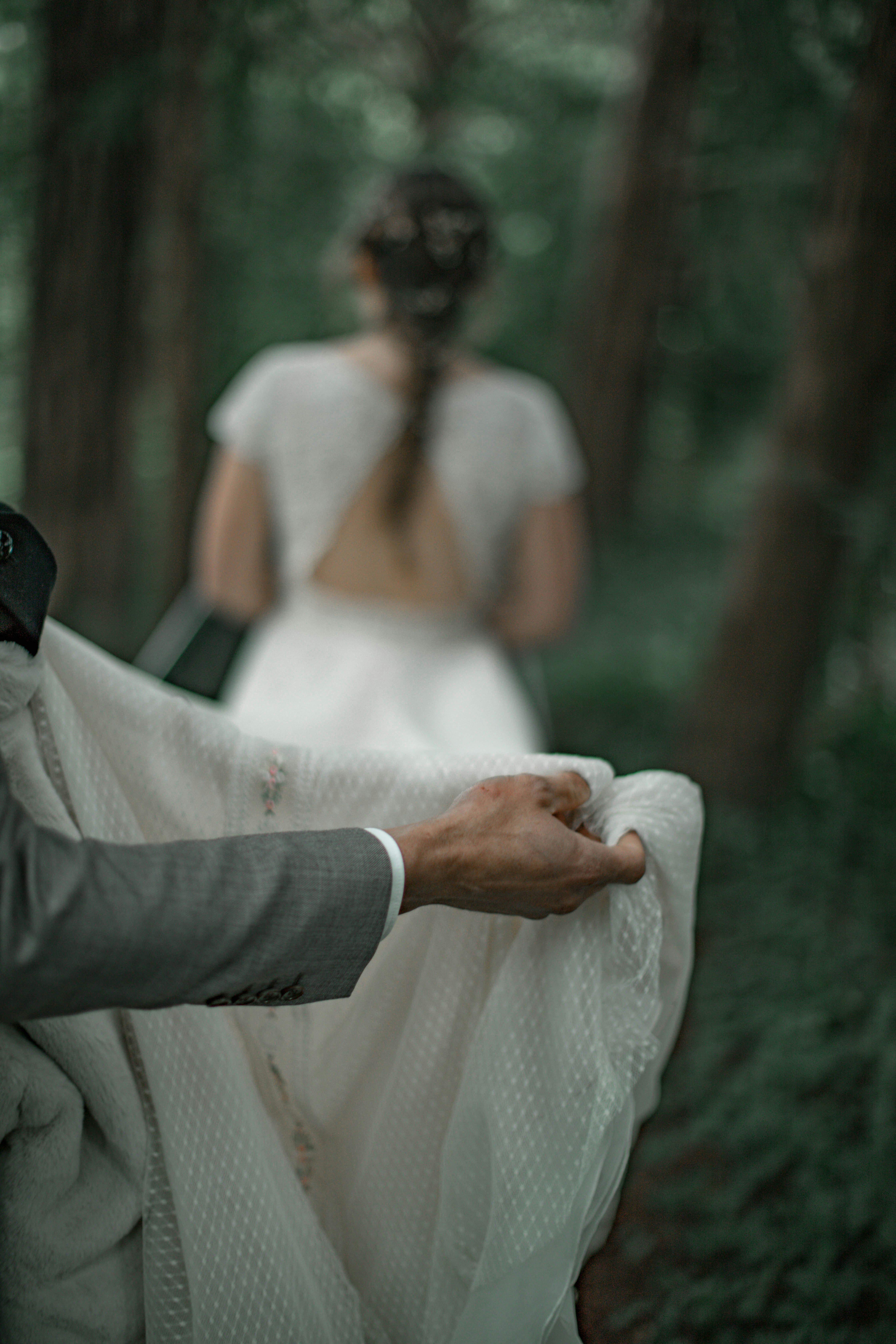 A groom gently holds the bride's dress as they walk through a lush green forest, capturing a moment of intimacy and connection.