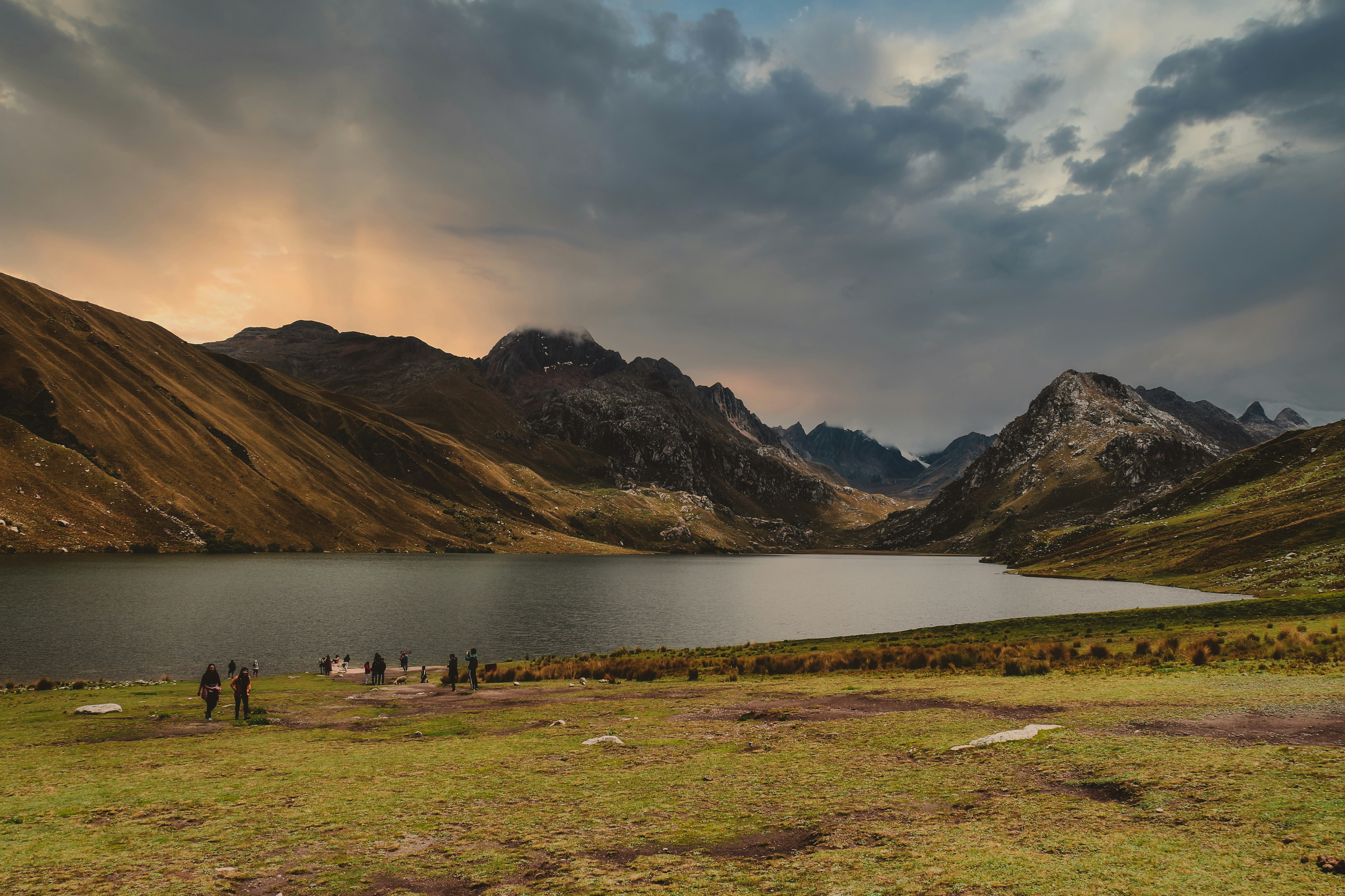 A tranquil lakeside scene framed by majestic mountains under a dramatic sky, with silhouettes of people enjoying the view.