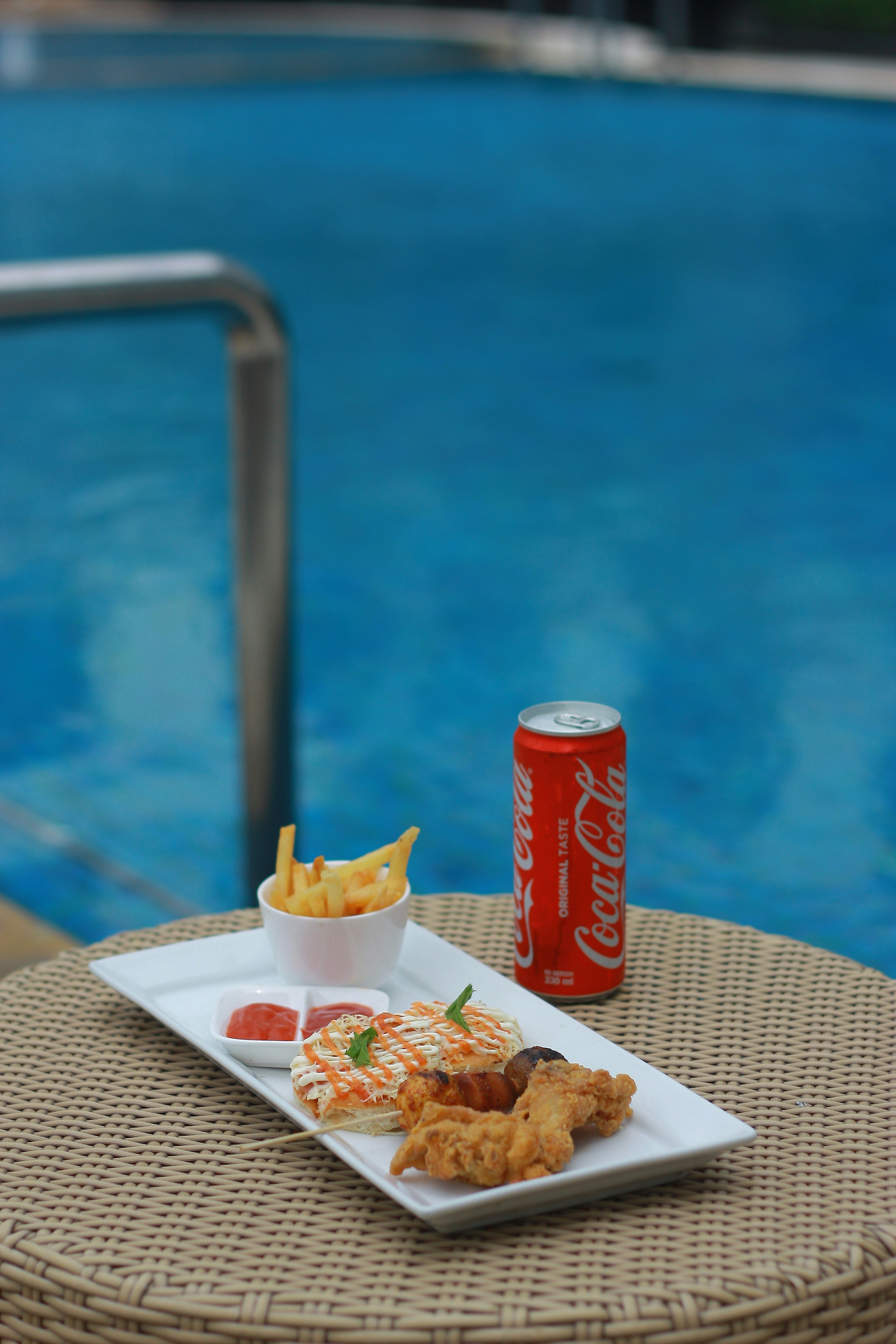 Plate of fried chicken, fries, and sauces accompanied by a Coca-Cola can, set against a tranquil blue pool backdrop.