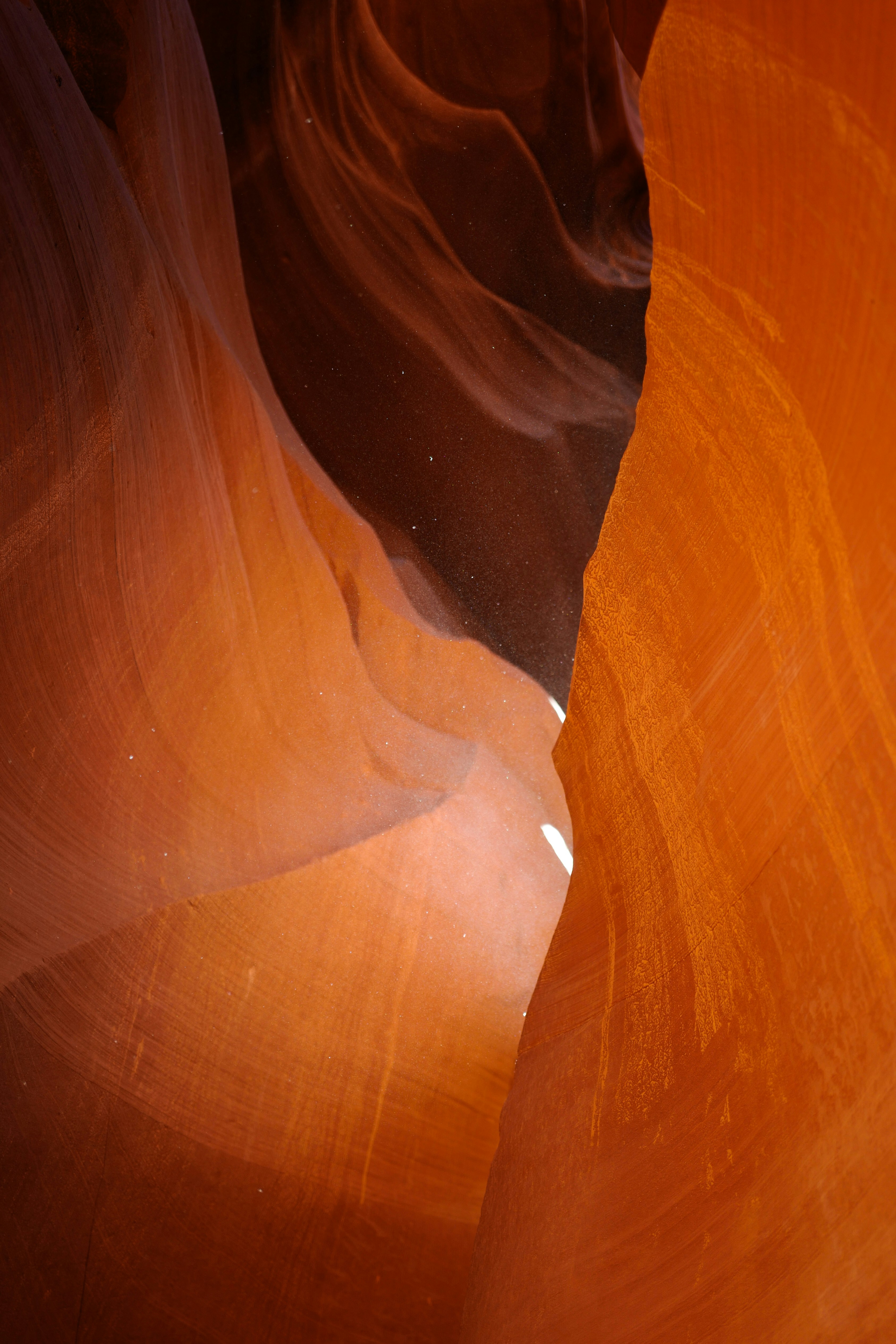 A photograph of a slot canyon bathed in warm orange sandstone, with a narrow opening at the lower center letting in bright light. Dust motes drift in the beam, emphasizing the canyon's sculpted curves.