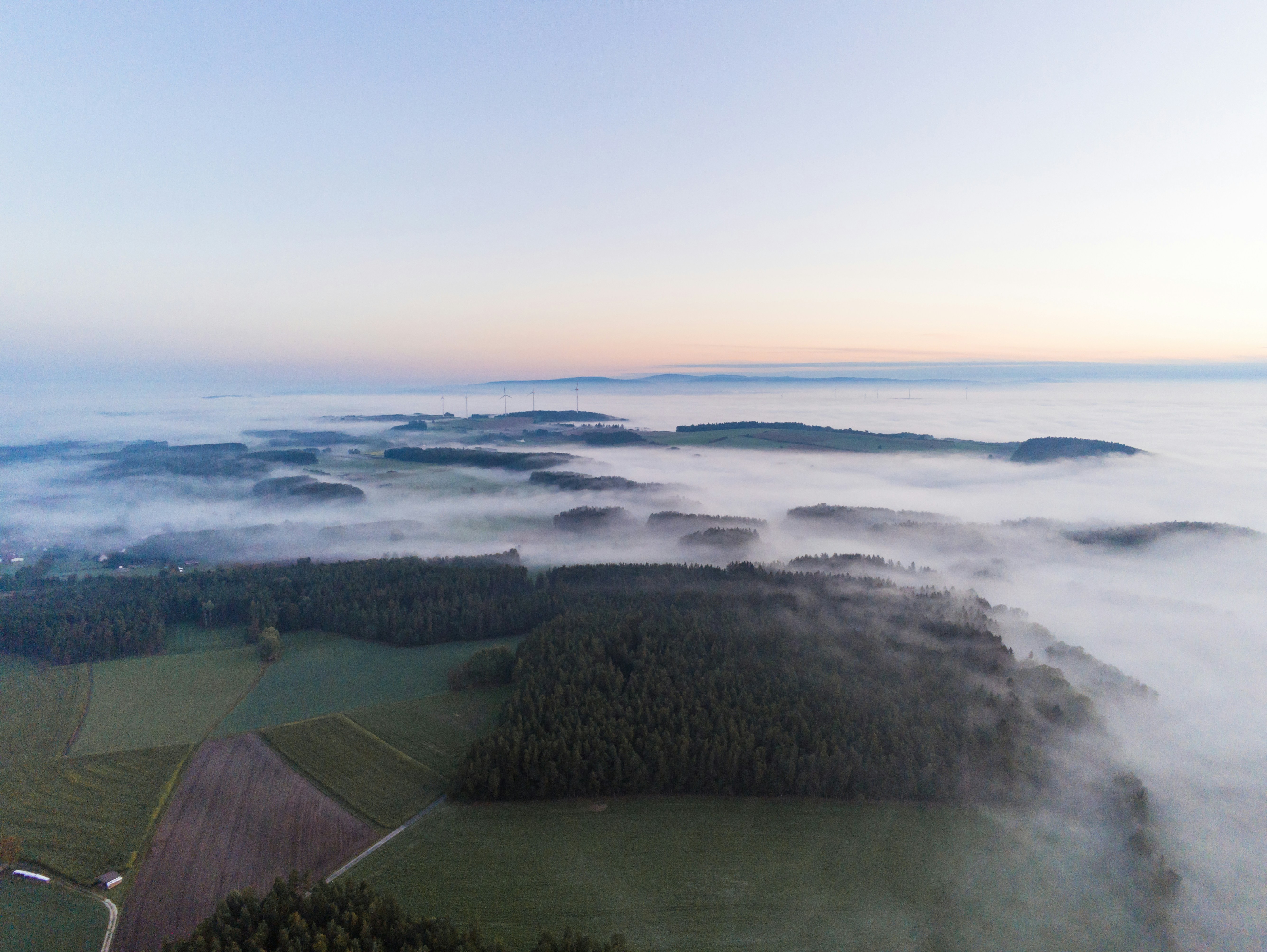 Aerial view of fog enveloping forested hills and patchwork fields at sunrise.