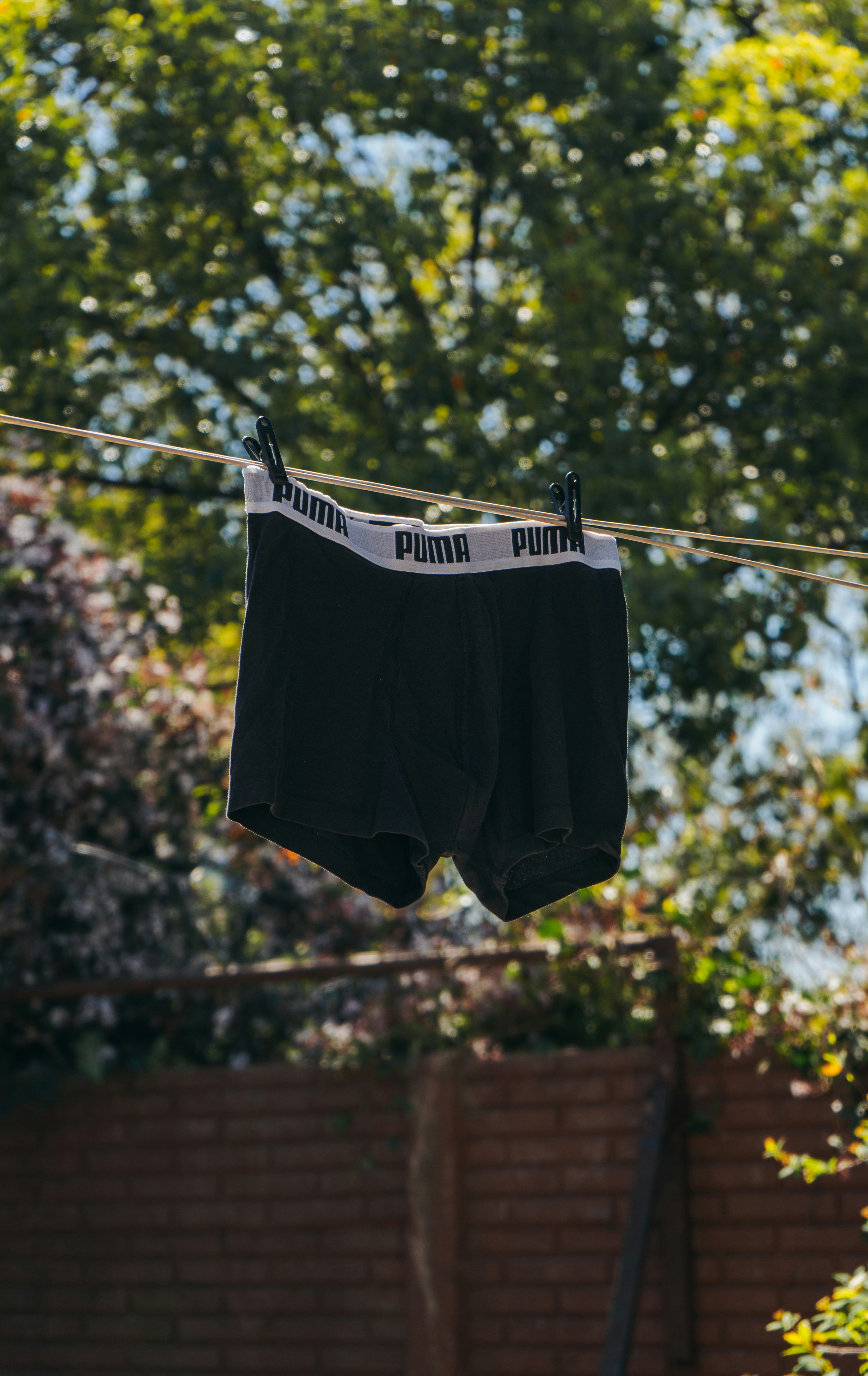 Black boxer shorts hanging on a clothesline against a backdrop of lush greenery and brick wall.