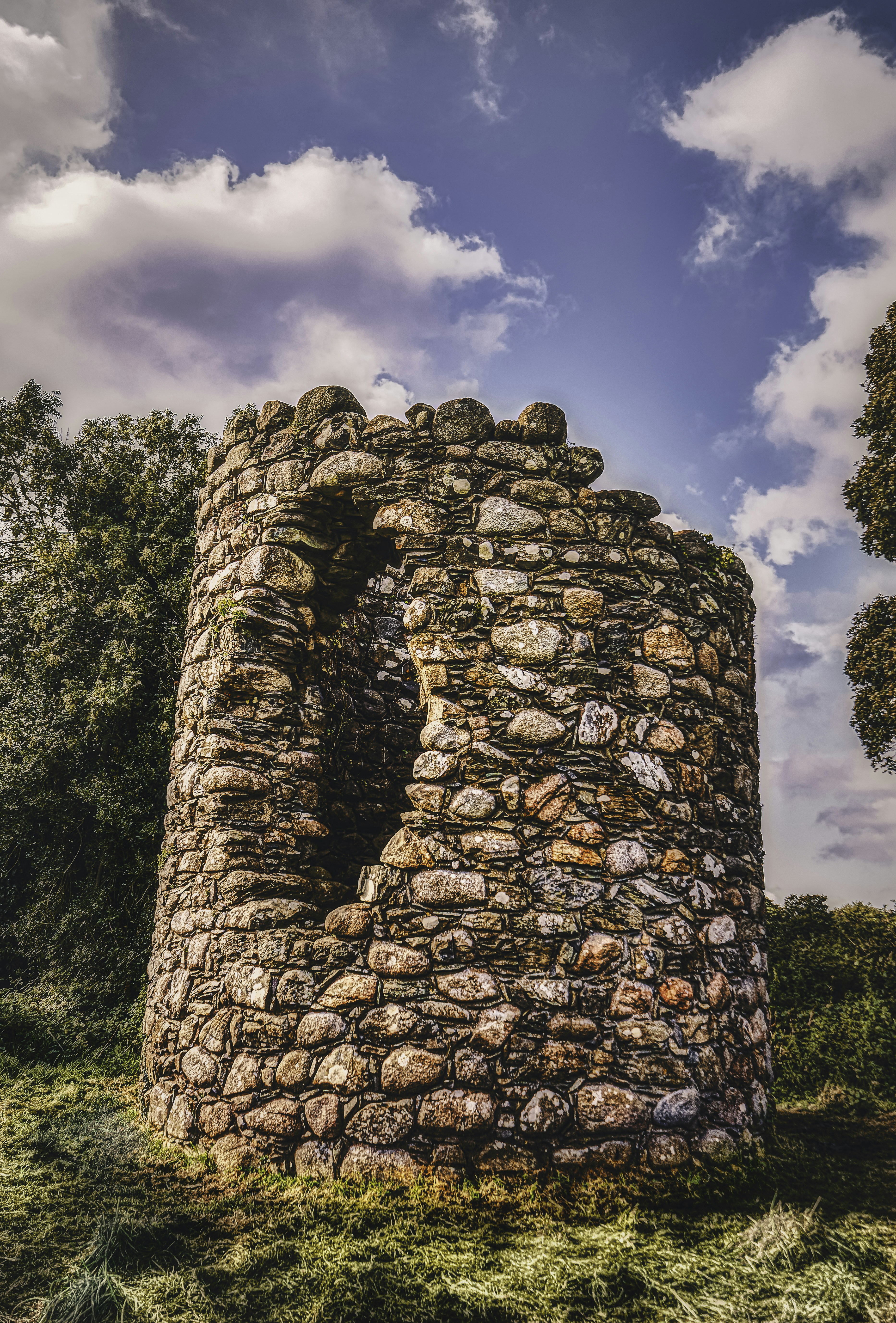 Ancient stone tower partially covered in greenery against a dramatic sky, showcasing its weathered texture and historical significance.