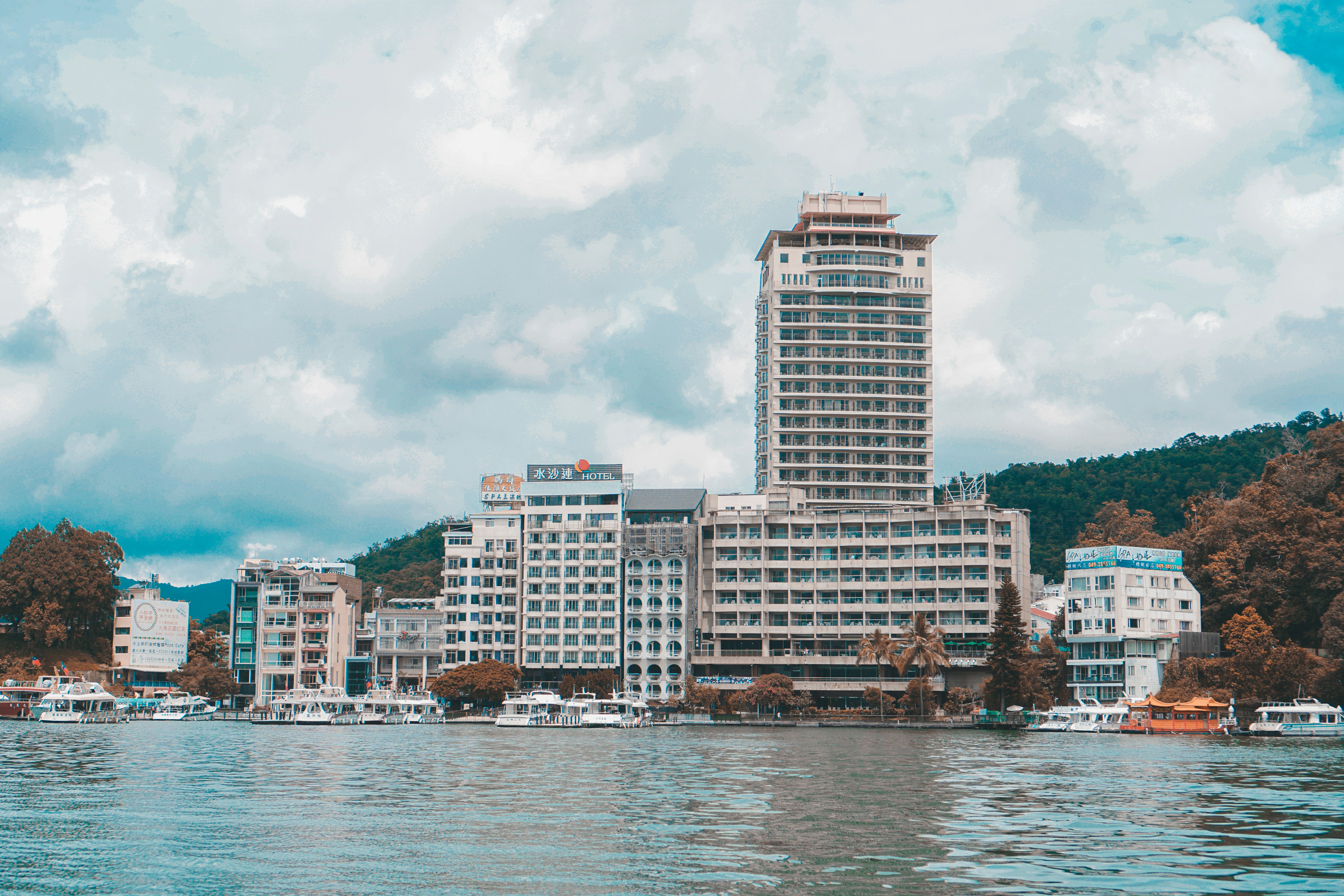Tall buildings with a backdrop of lush hills and a reflective lake under a cloudy sky.