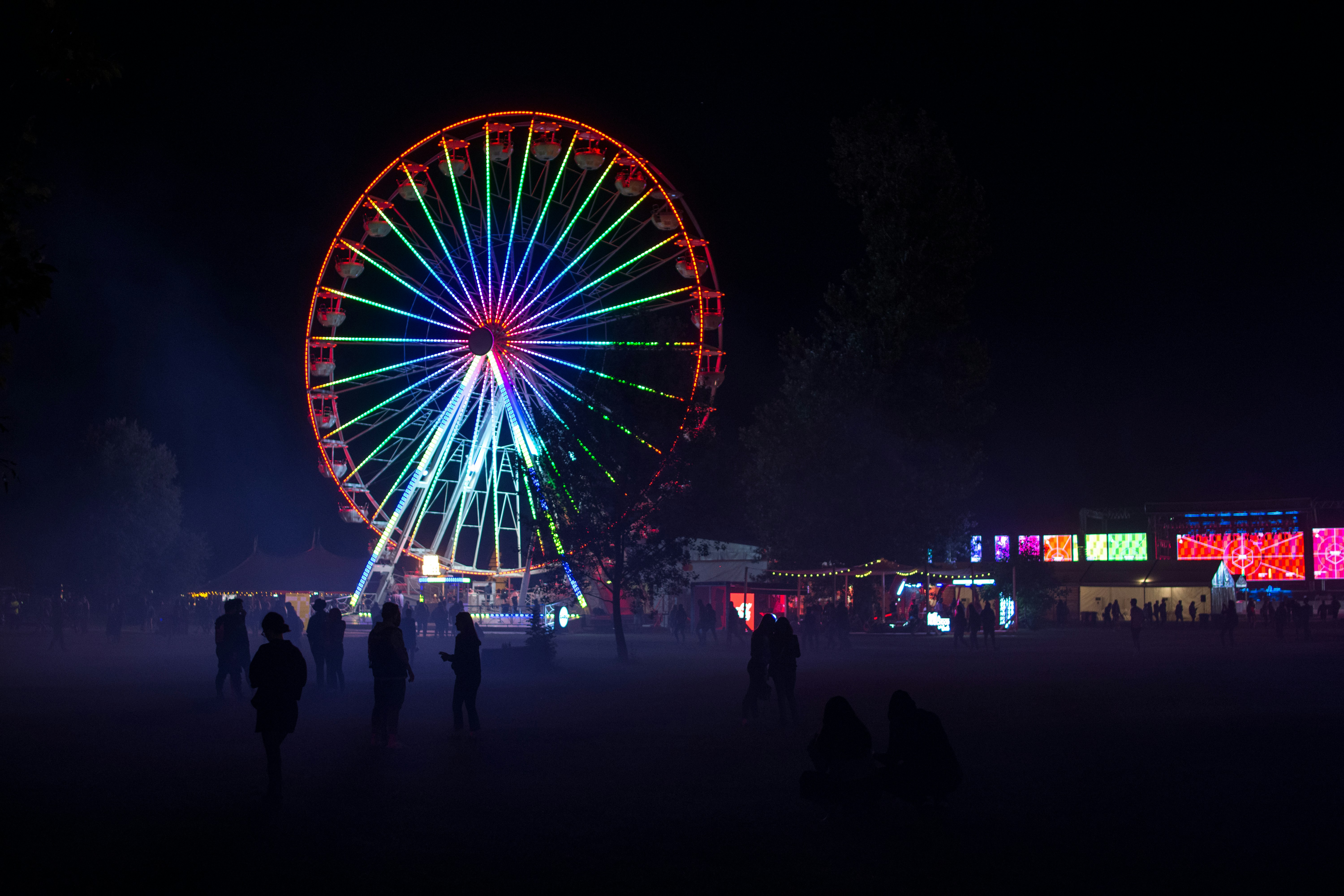 Silhouetted figures stroll through a misty park with a brightly lit Ferris wheel and colorful vendor stalls at night.
