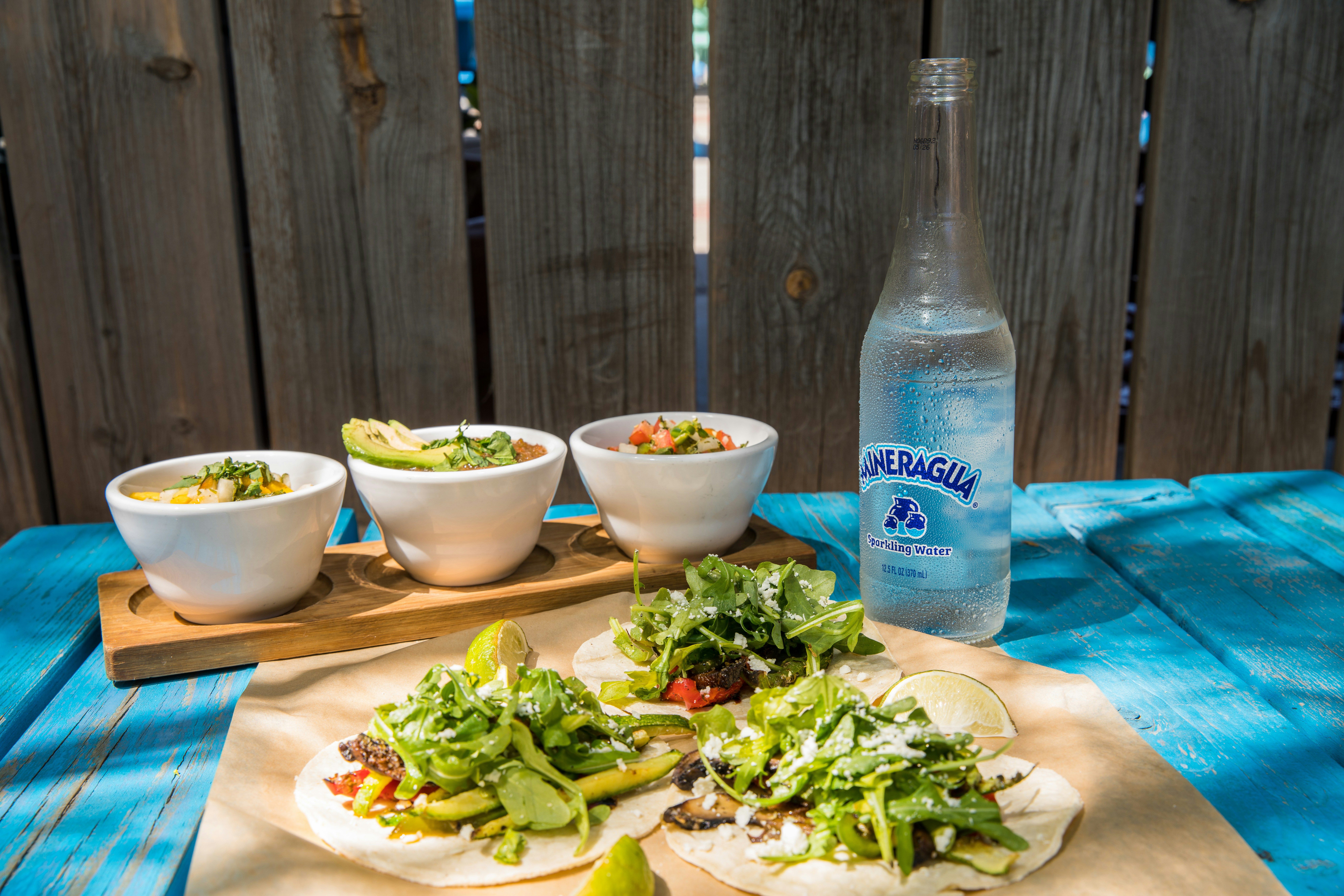 Two tacos topped with fresh greens and colorful ingredients, accompanied by a bottle of sparkling water and bowls of salsa, set against a rustic wooden backdrop.