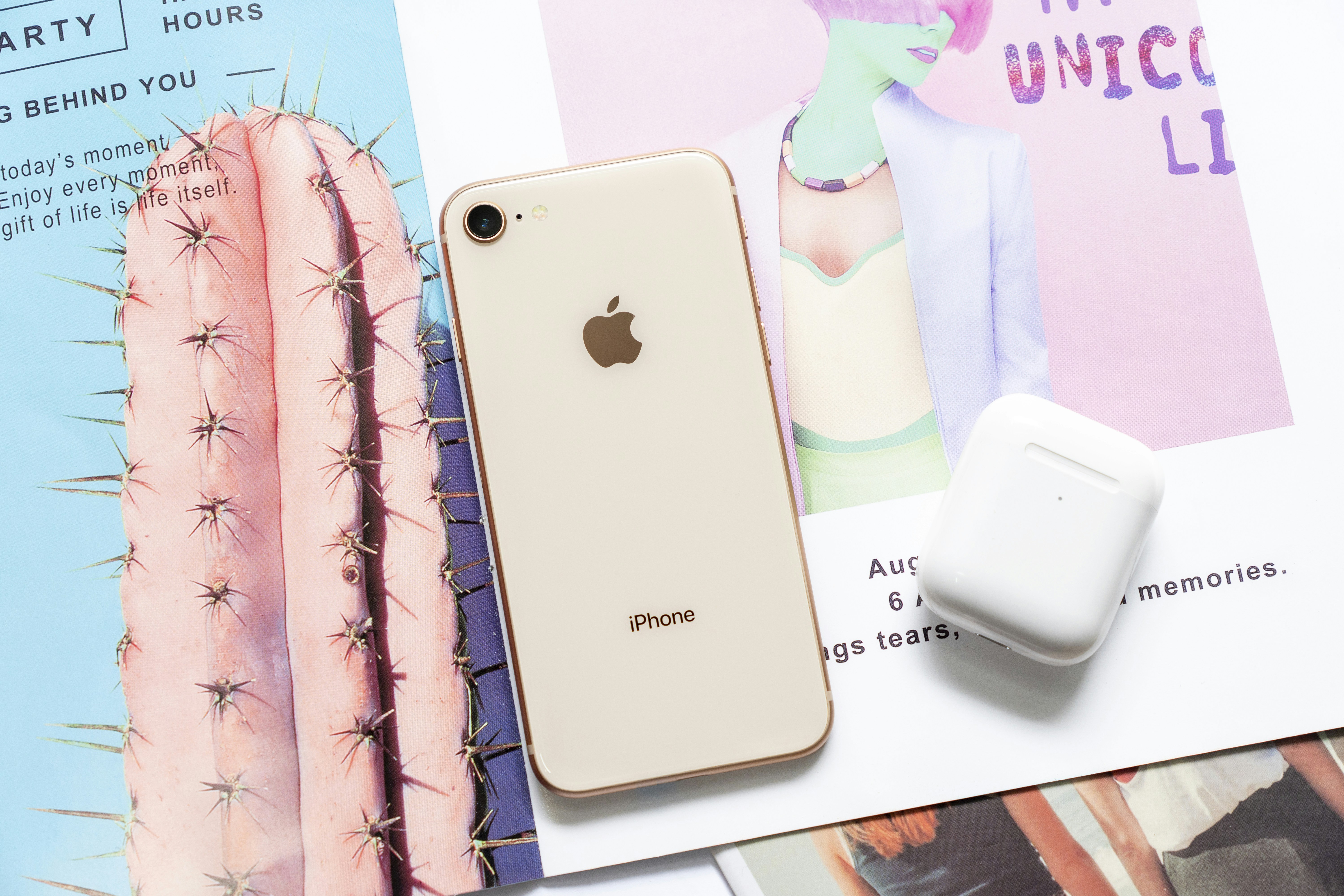 Gold iPhone and white AirPods case atop colorful, artistic background featuring a cactus illustration.