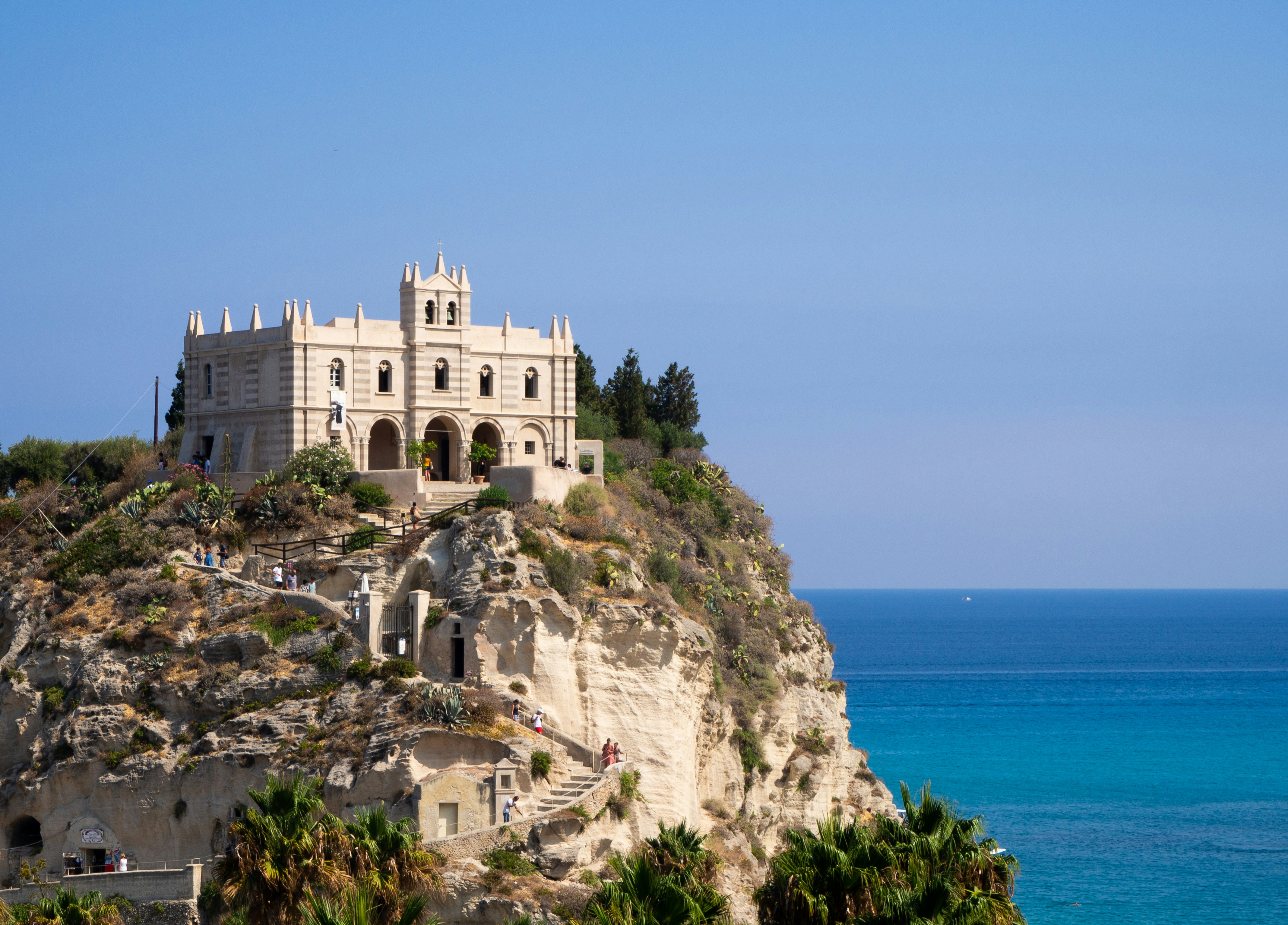 Historic building perched on a rugged cliff above the turquoise sea under a clear blue sky.