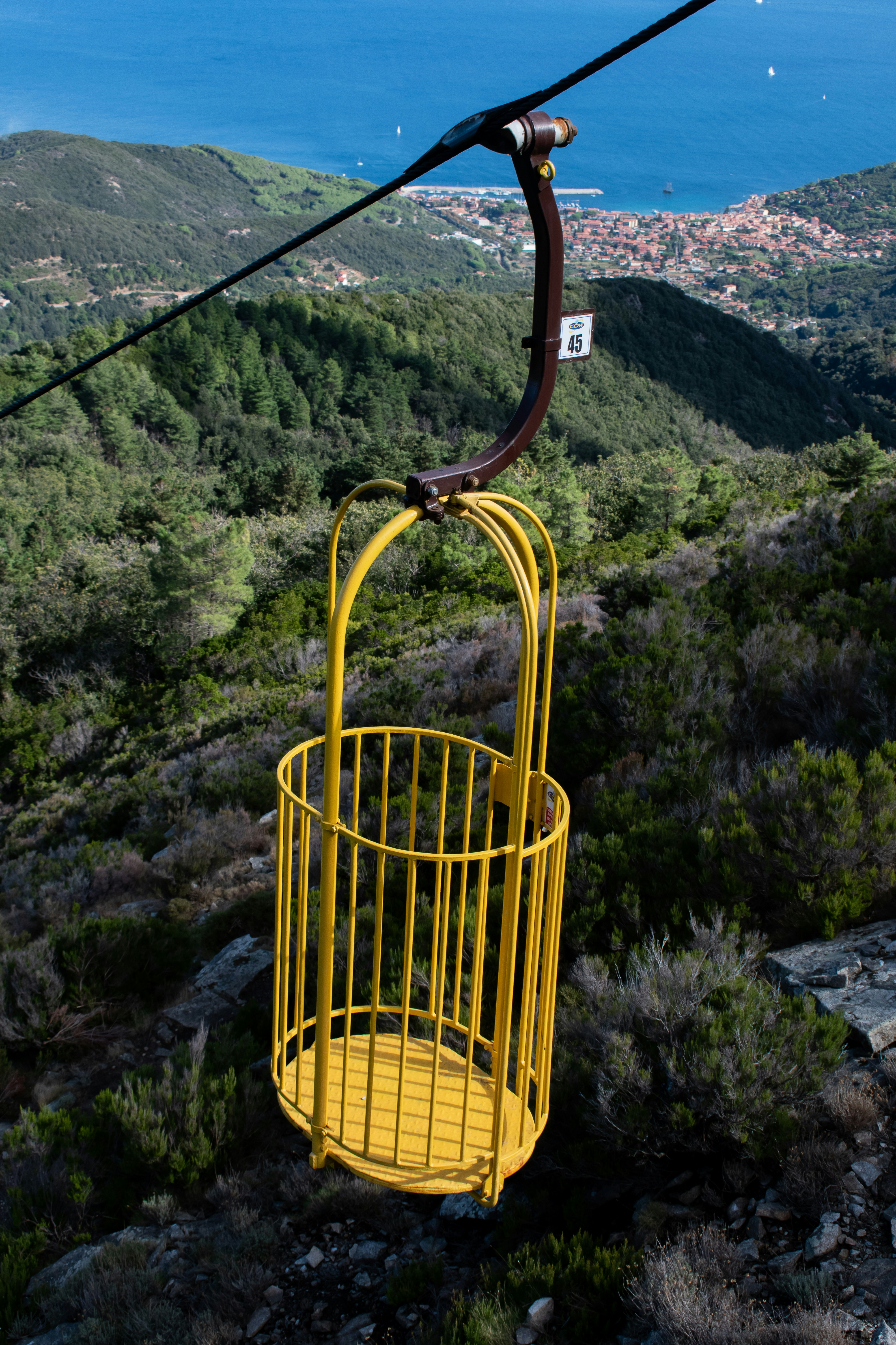 A yellow basket suspended from a cable overlooks a lush hillside and a coastal town below. The scene captures the essence of adventure and nature's beauty.