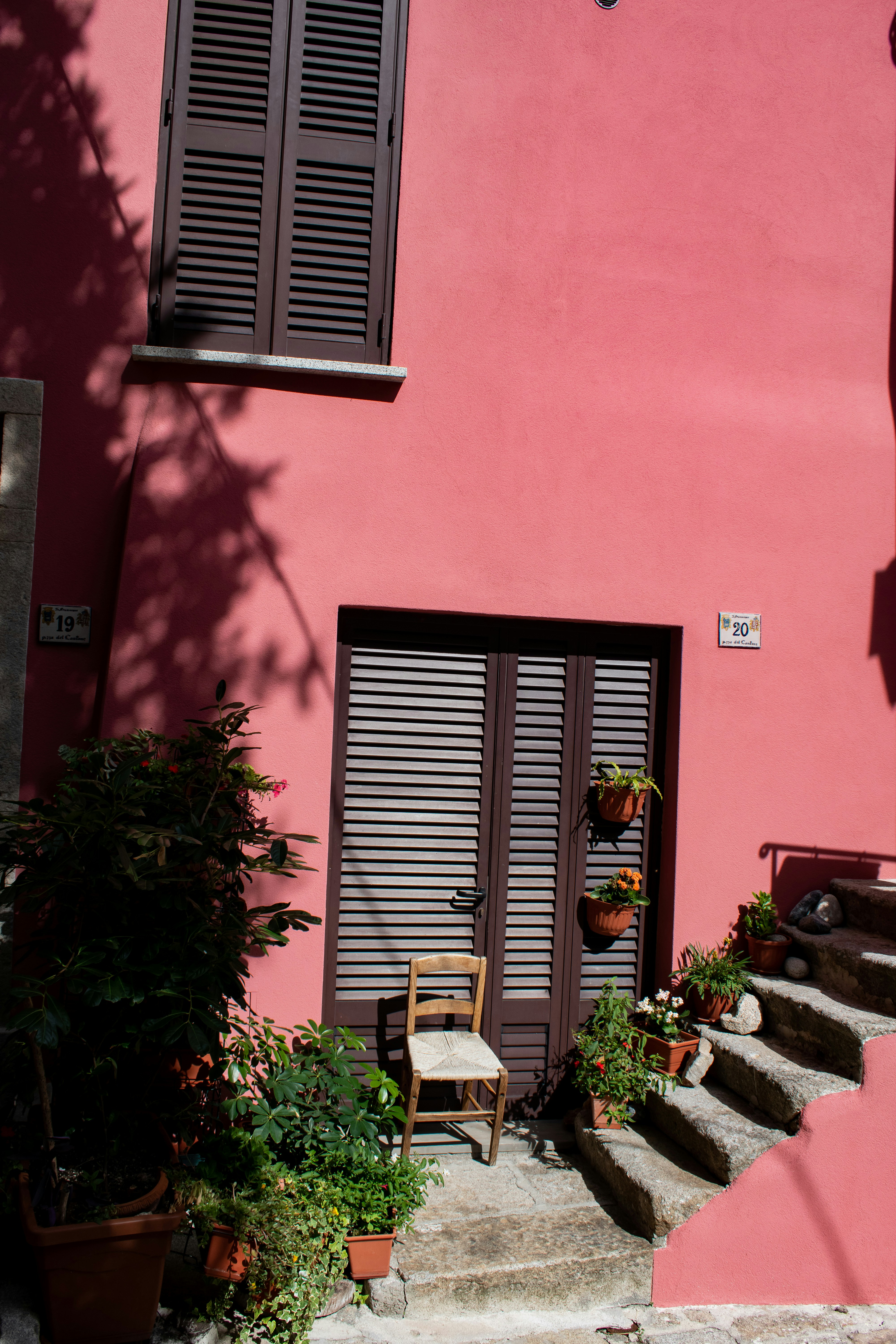 A quaint pink house adorned with potted plants and a wooden chair, inviting serenity and warmth. The interplay of light and shadow enhances the cozy atmosphere.