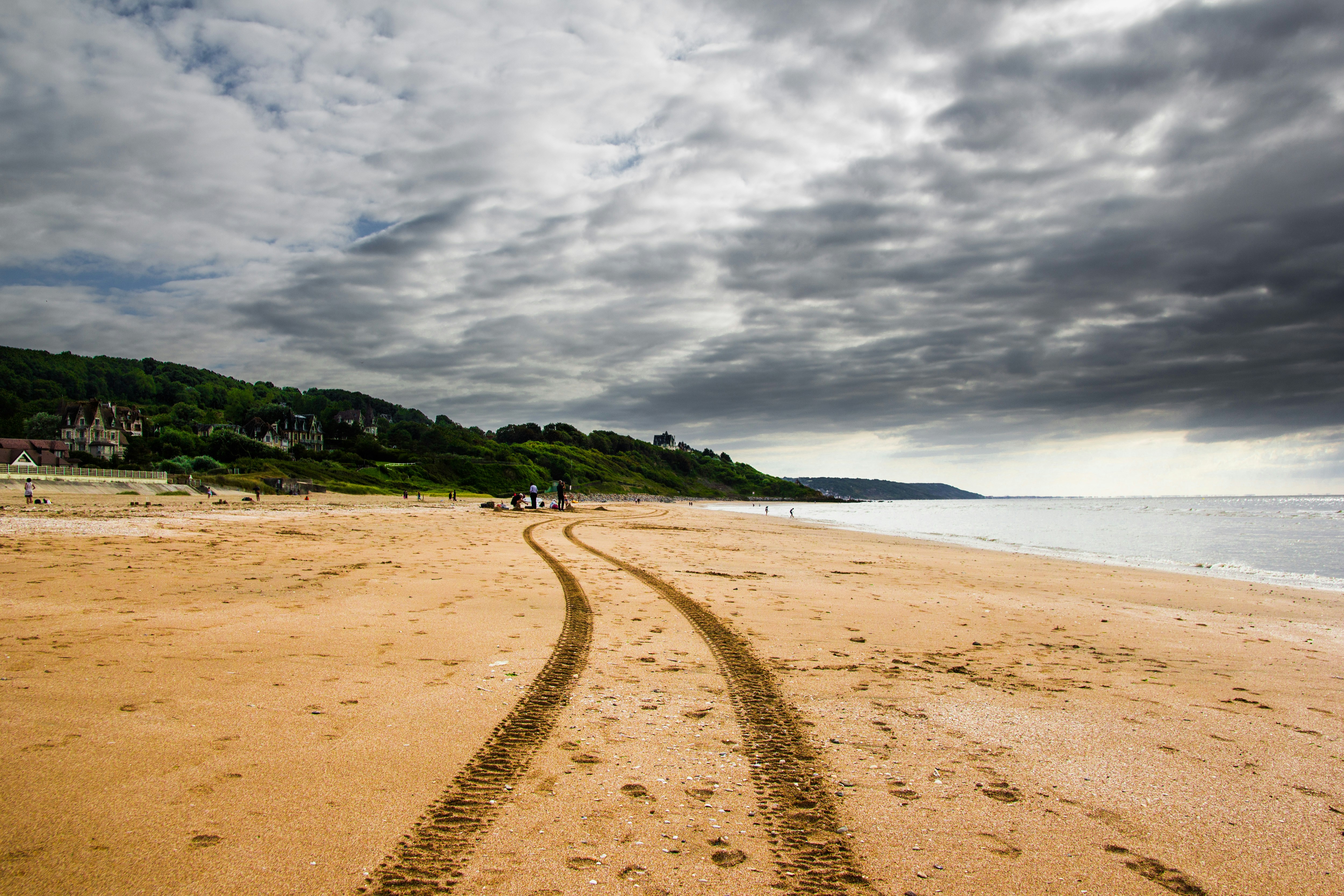 Tire tracks lead across a sandy beach under a dramatic sky, with distant figures enjoying the shoreline. The scene captures a tranquil moment by the sea.