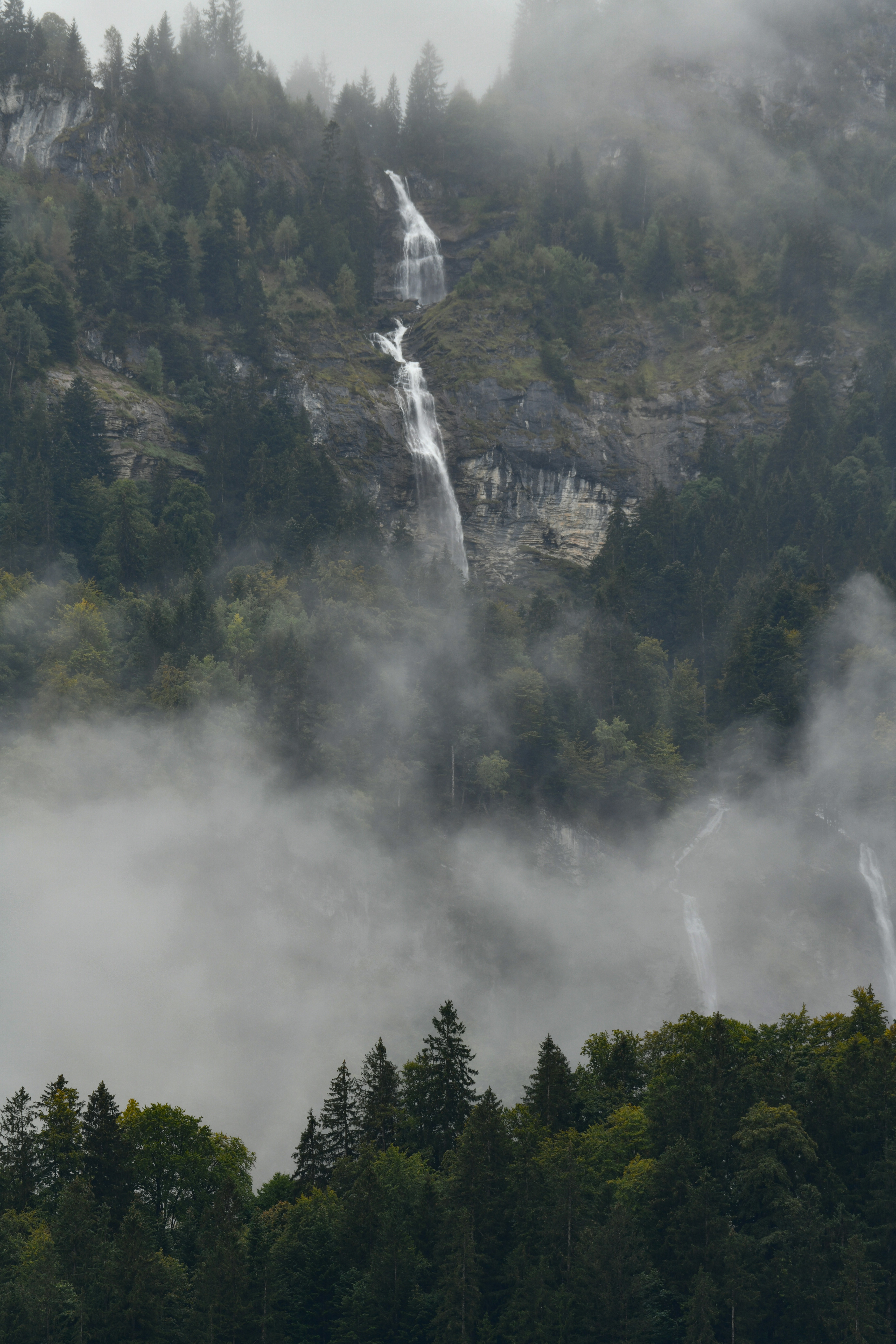 Waterfalls cascading down a rocky cliff, partially obscured by mist and surrounded by lush green trees.