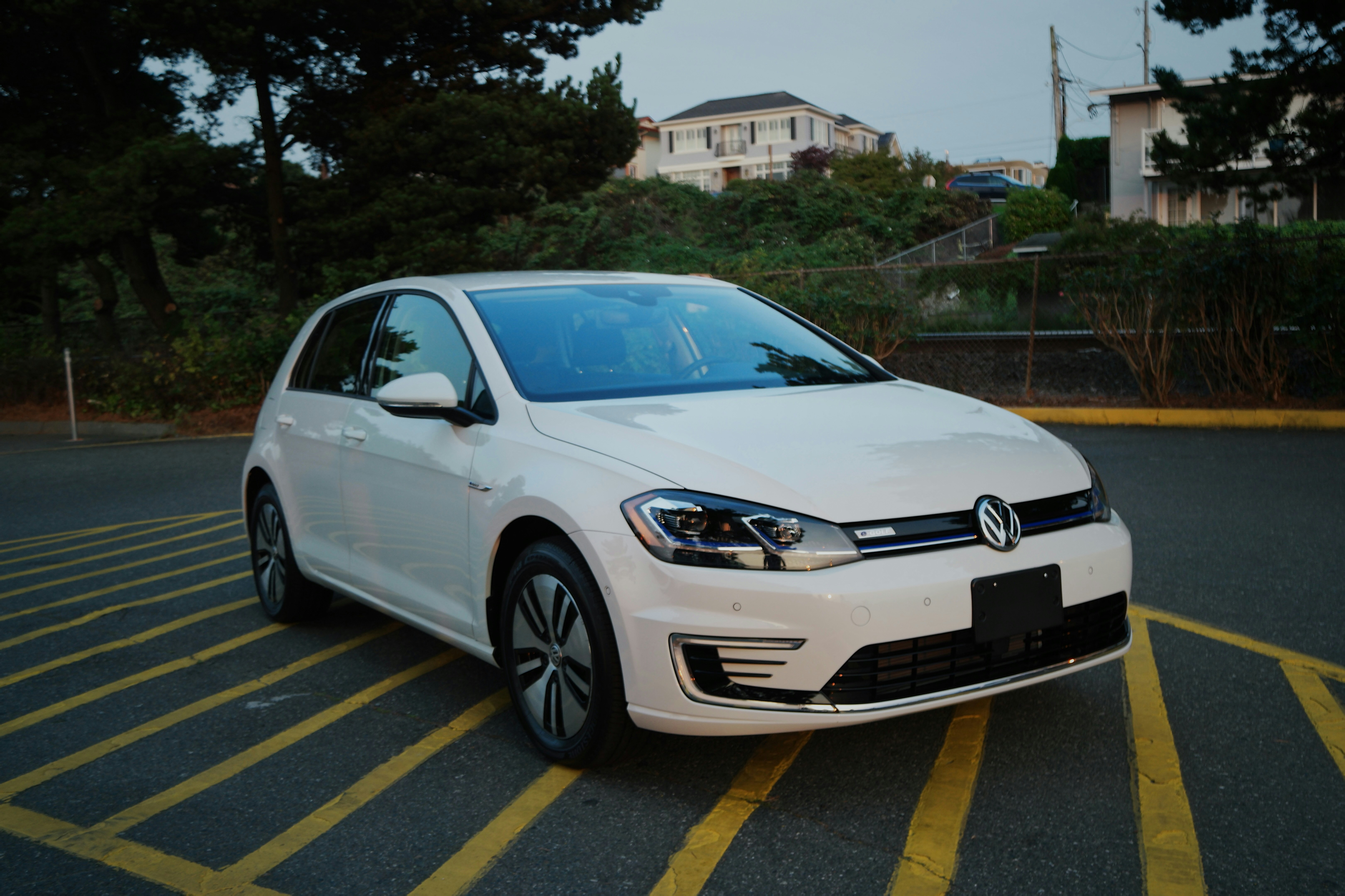 White Volkswagen hatchback parked on a marked road with trees and buildings in the background.