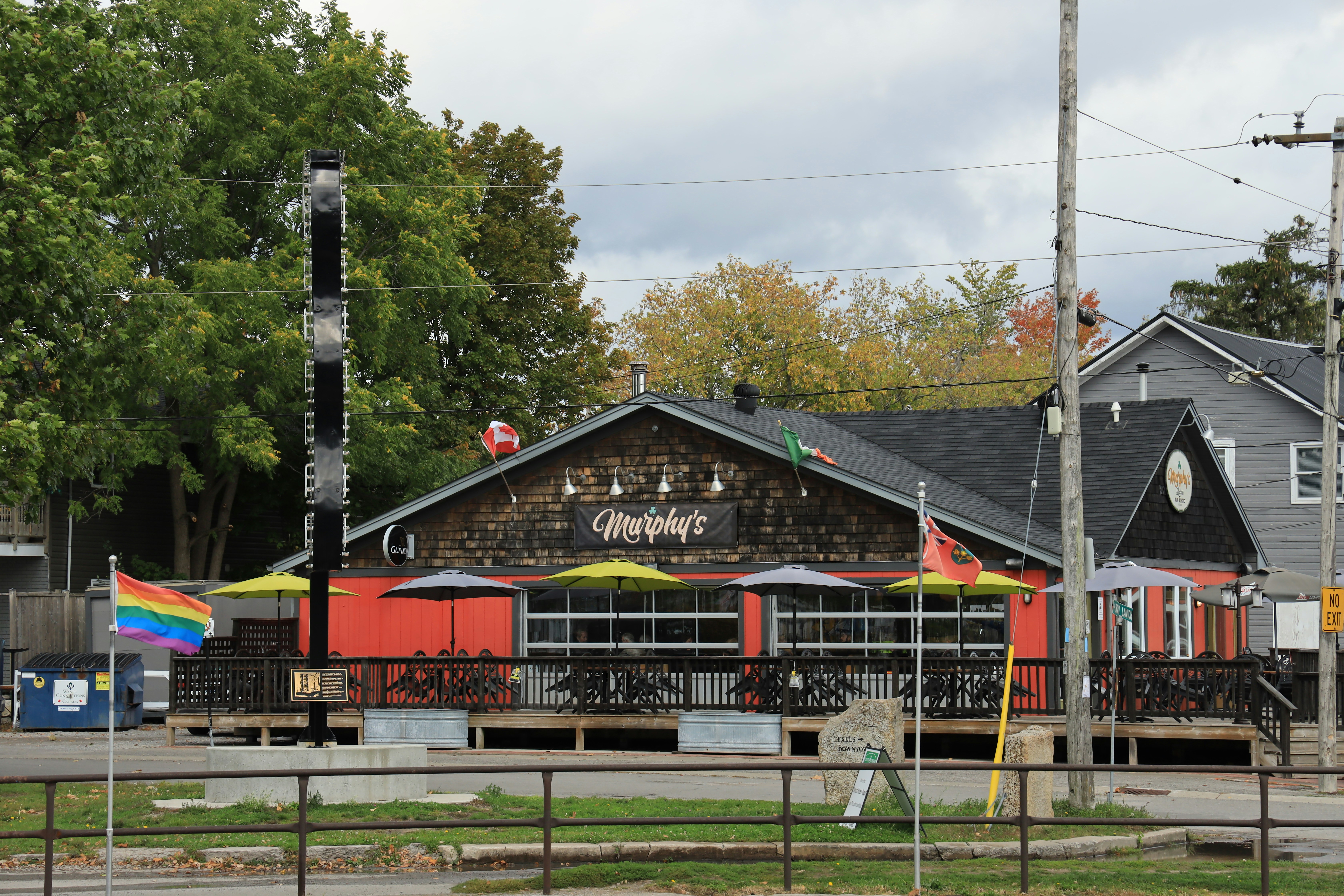 Colorful flags flutter in front of a lively restaurant with outdoor seating, framed by autumn foliage. The inviting atmosphere encourages social gatherings.