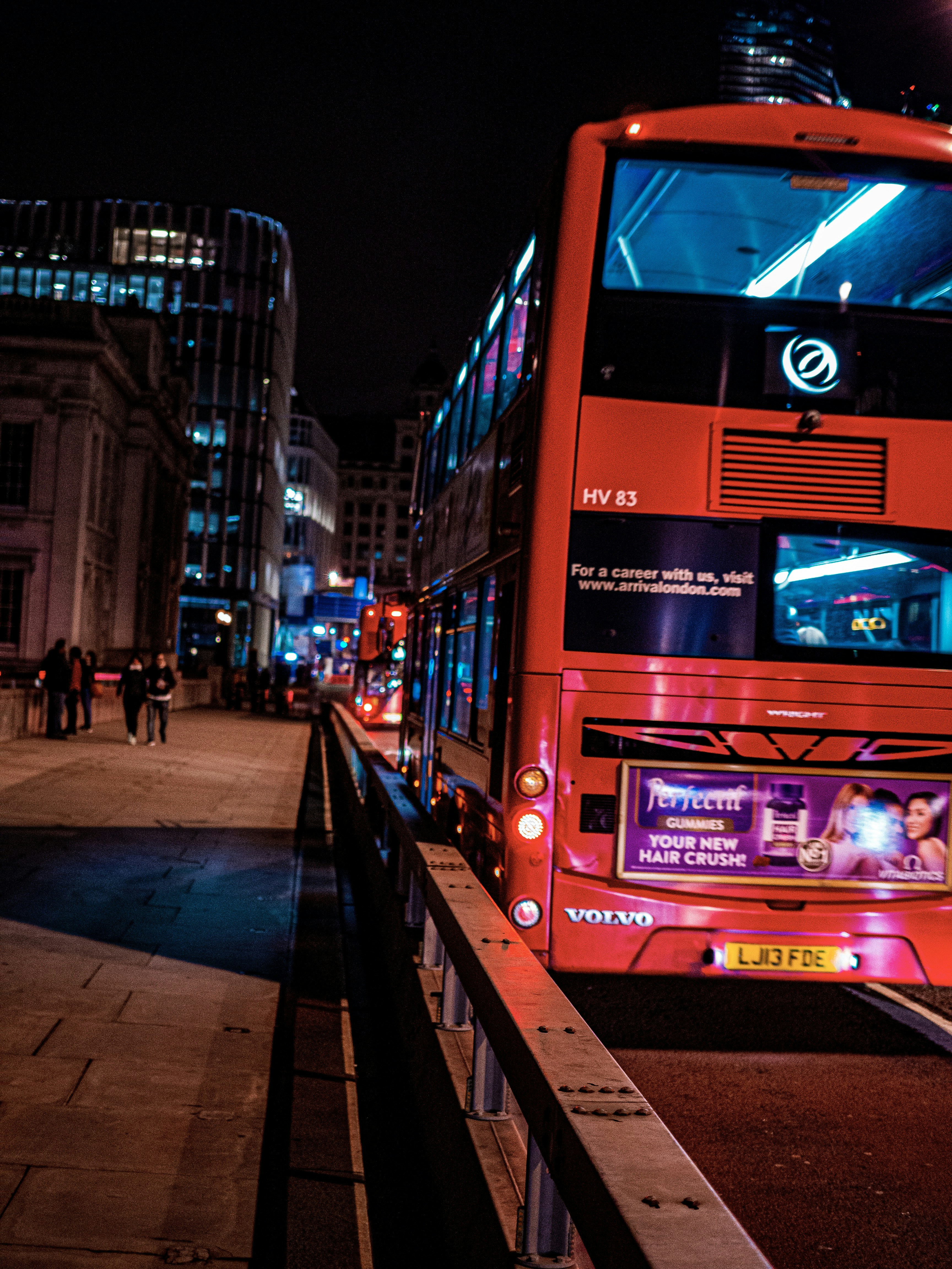 A row of illuminated red buses lined up on a bustling city street at night, showcasing vibrant advertisements and urban architecture.
