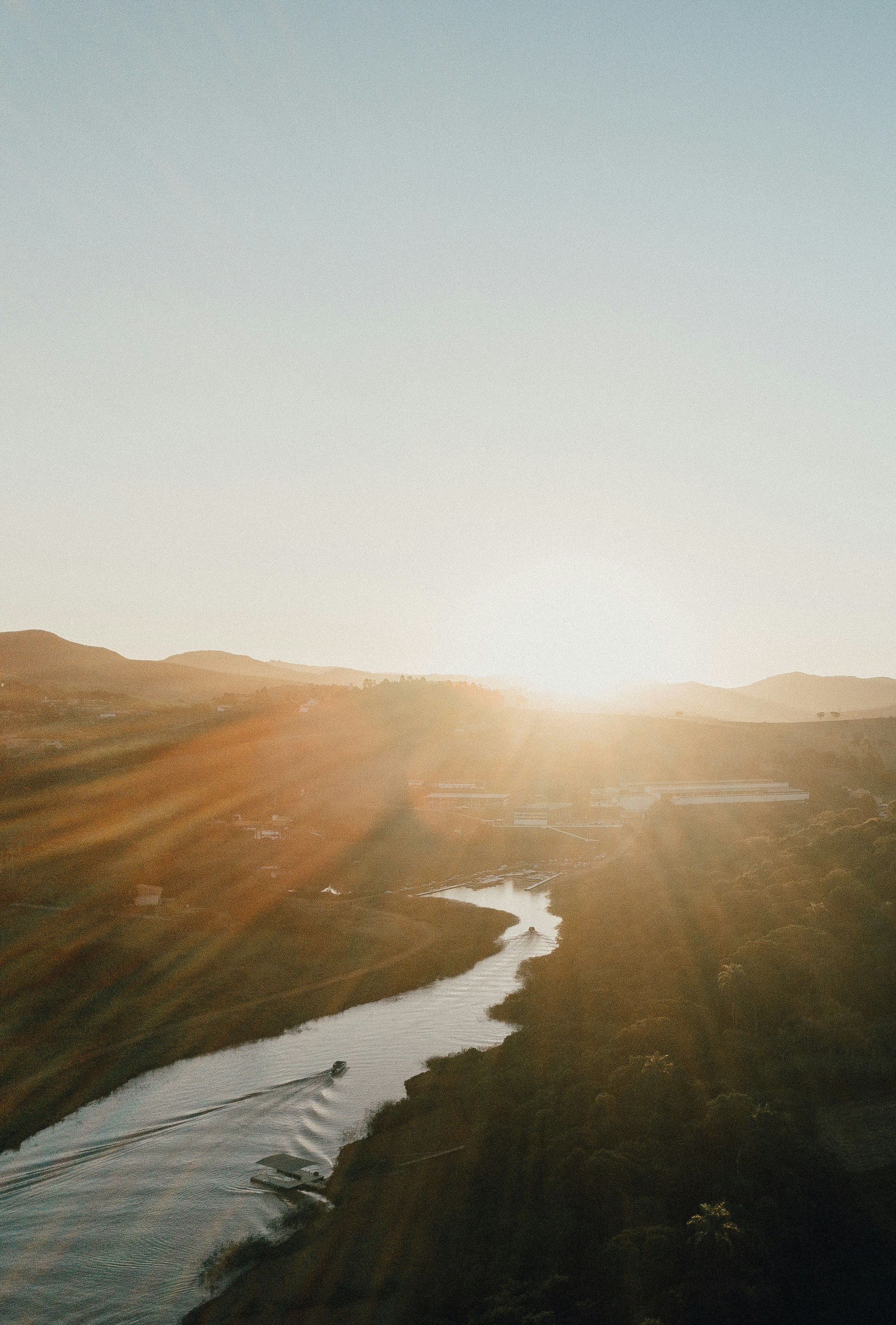 A tranquil river winding through a lush landscape under a setting sun, with rays illuminating the scene. The image captures the serene beauty of nature's transition from day to night.