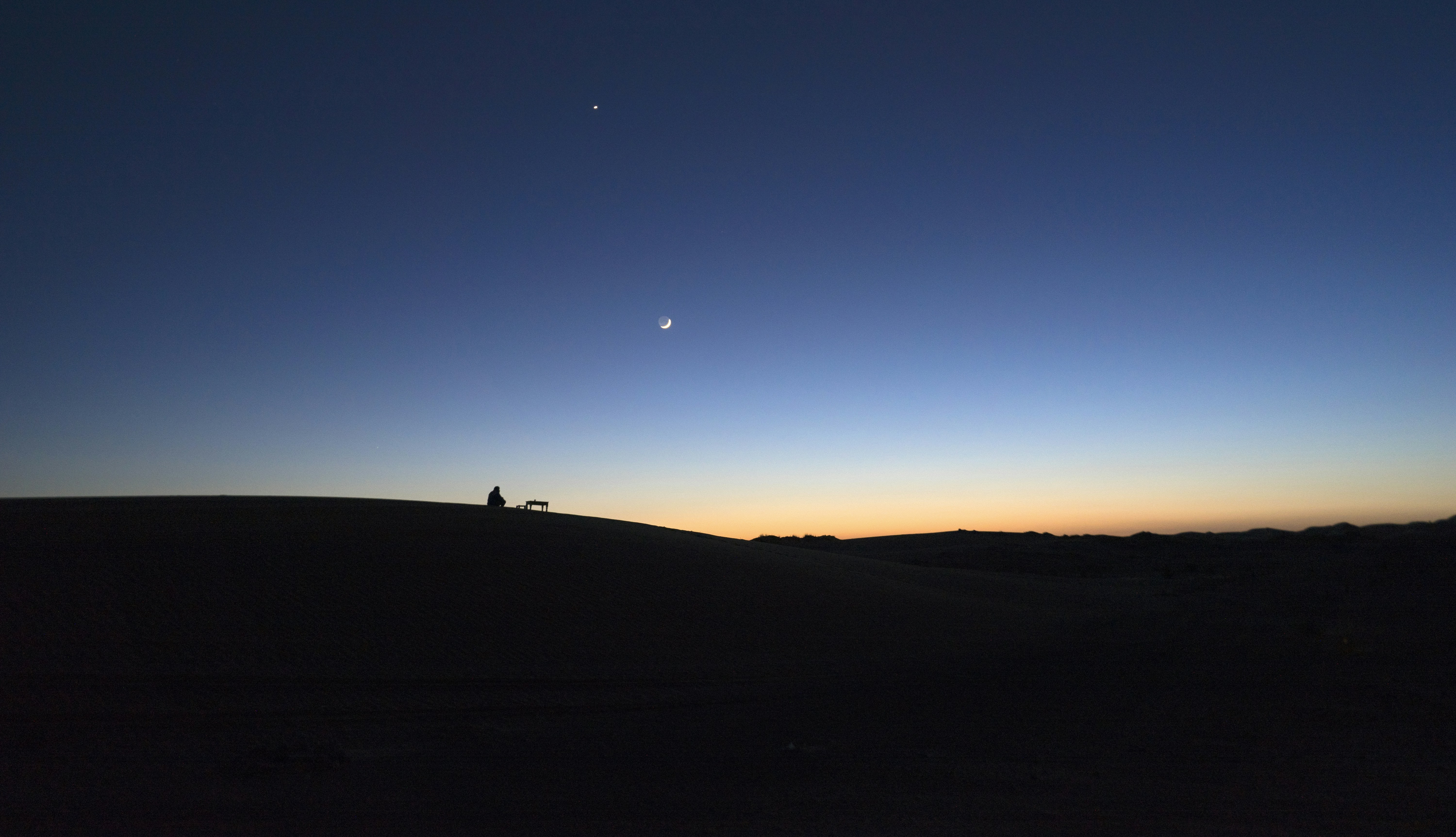 A crescent moon and one of the planets appear in the night sky as a man meditates, watching the final minutes of the day.
