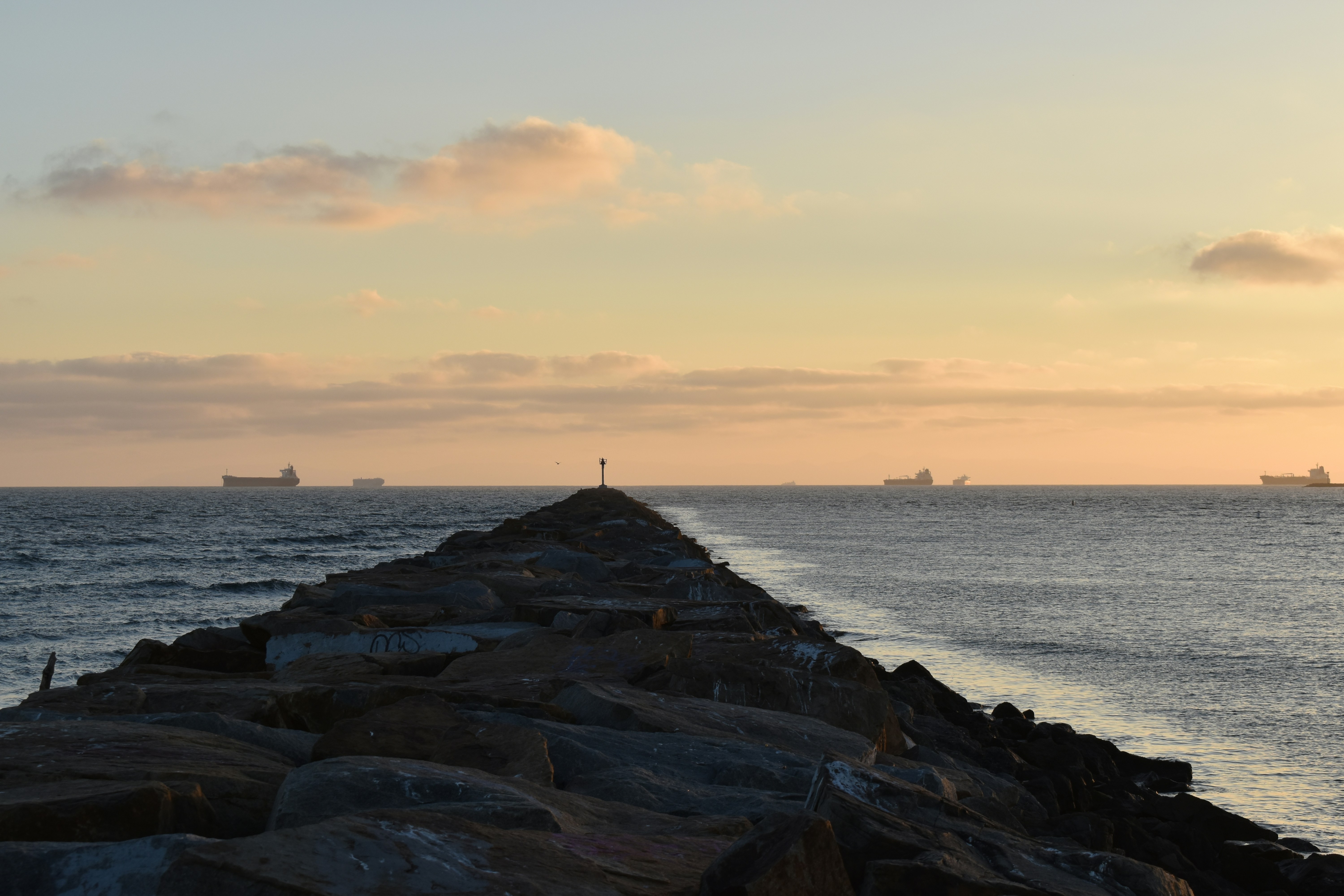 Rocky jetty extending into the ocean under a warm sunset sky with distant ships on the horizon.
