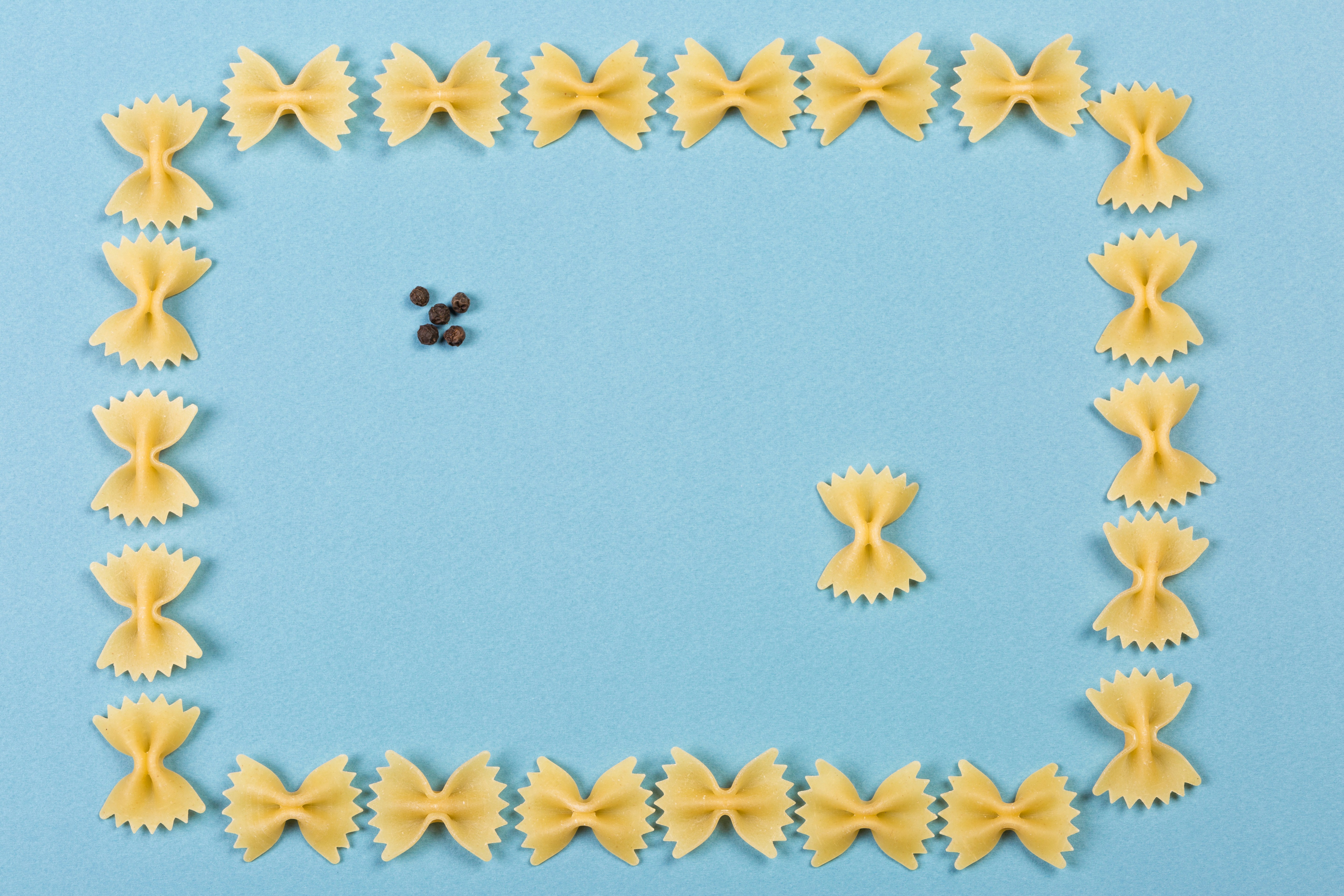 Farfalle pasta arranged in a rectangular frame on a blue background, with a few coffee beans placed inside. A playful culinary composition.