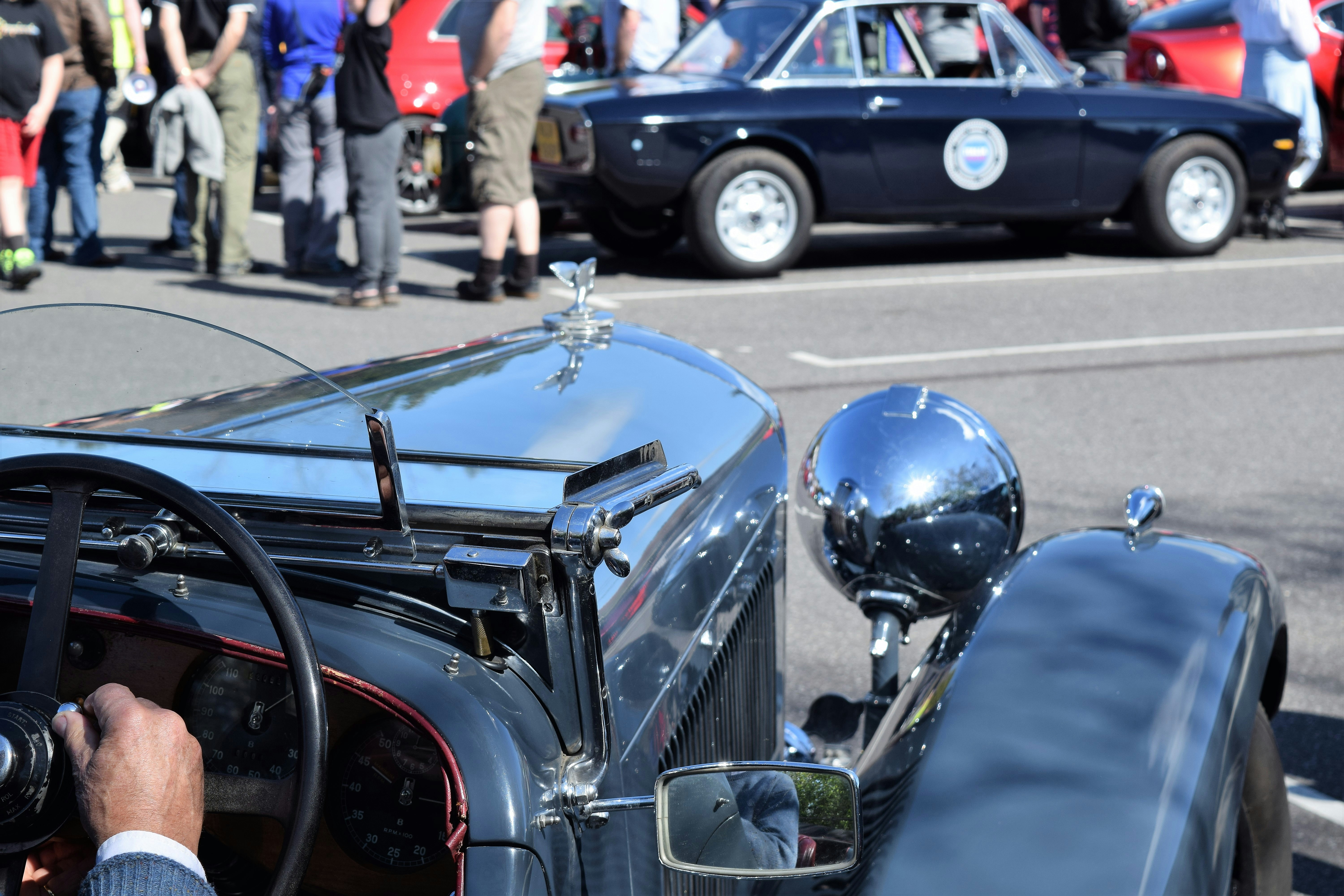 Classic car interior with a driver's hand on the steering wheel, showcasing vintage design elements, while modern vehicles and spectators are visible in the background.