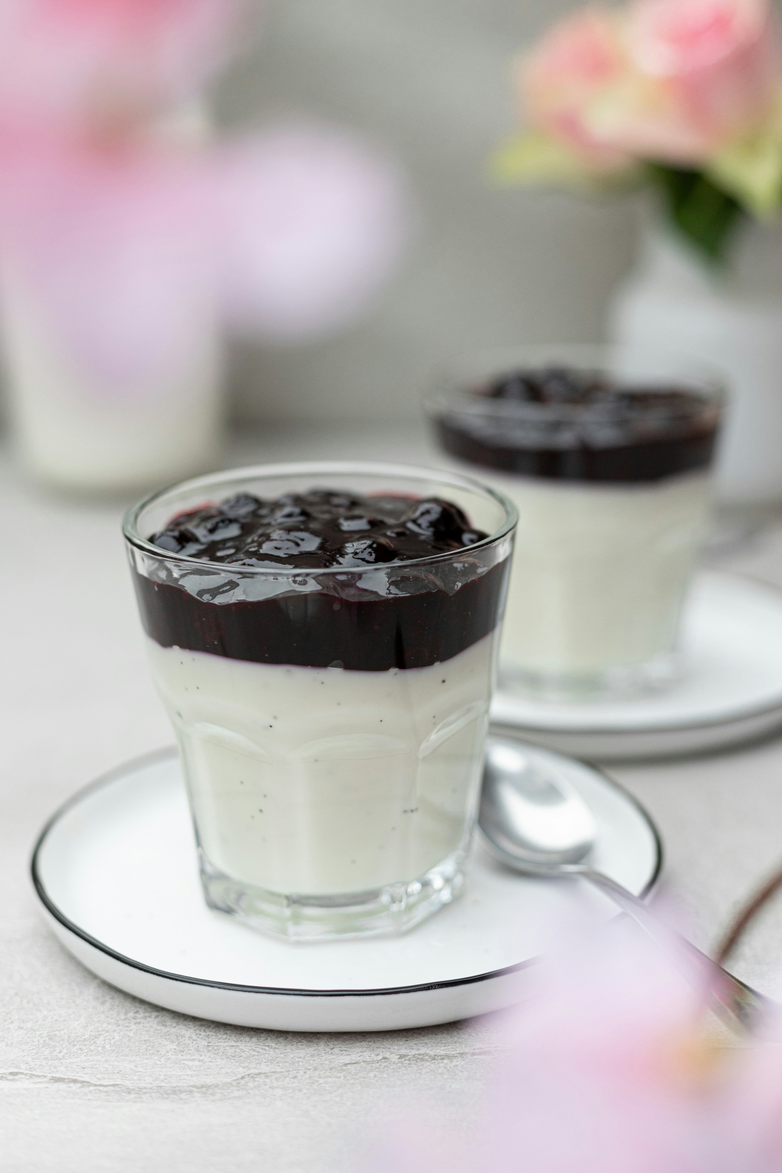 Creamy panna cotta topped with seasoned blueberry compote on a white plate, accompanied by soft pink flowers.