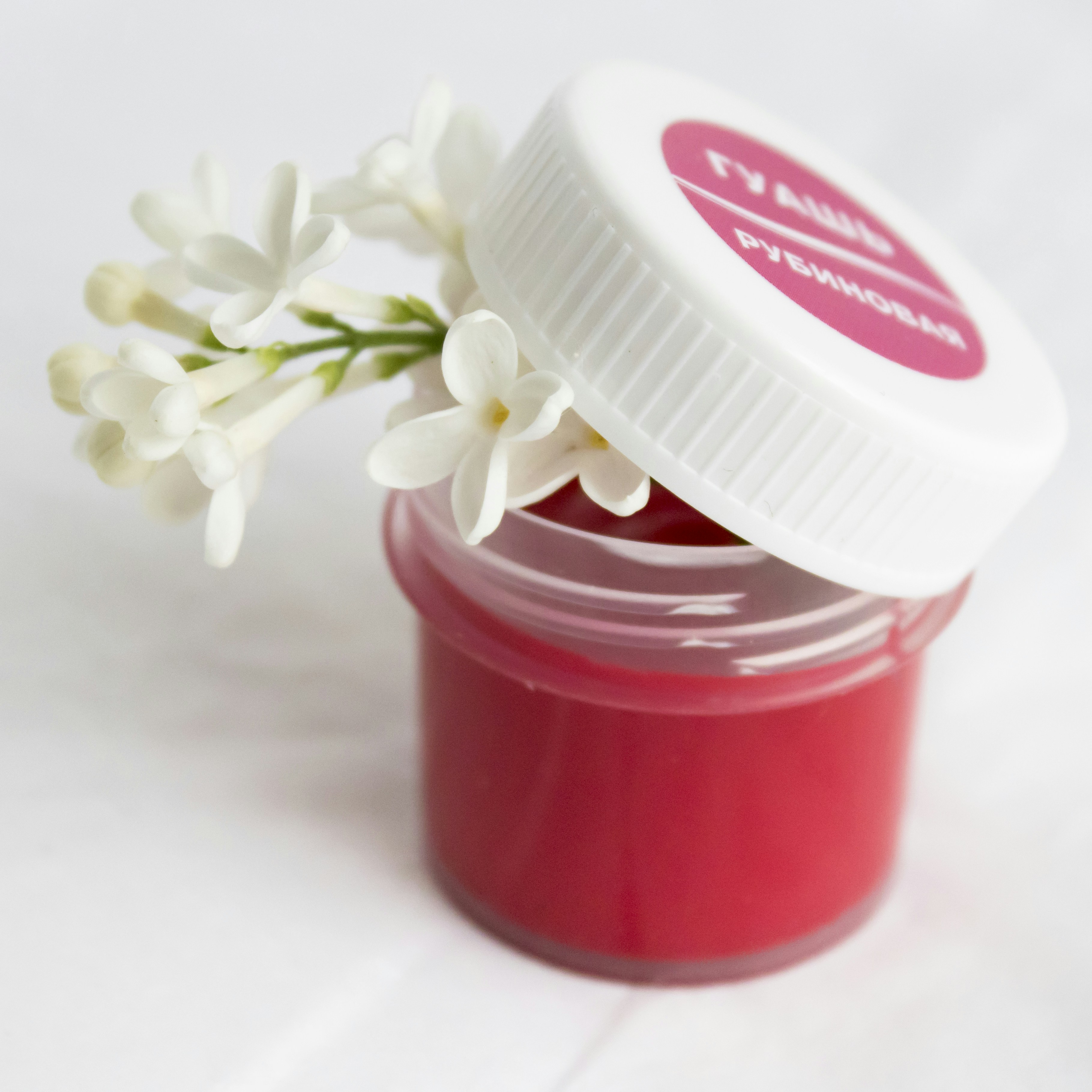 A small red jar with a white lid sits at a slight tilt, adorned by delicate white flowers, creating a minimal still life focused on color and texture.