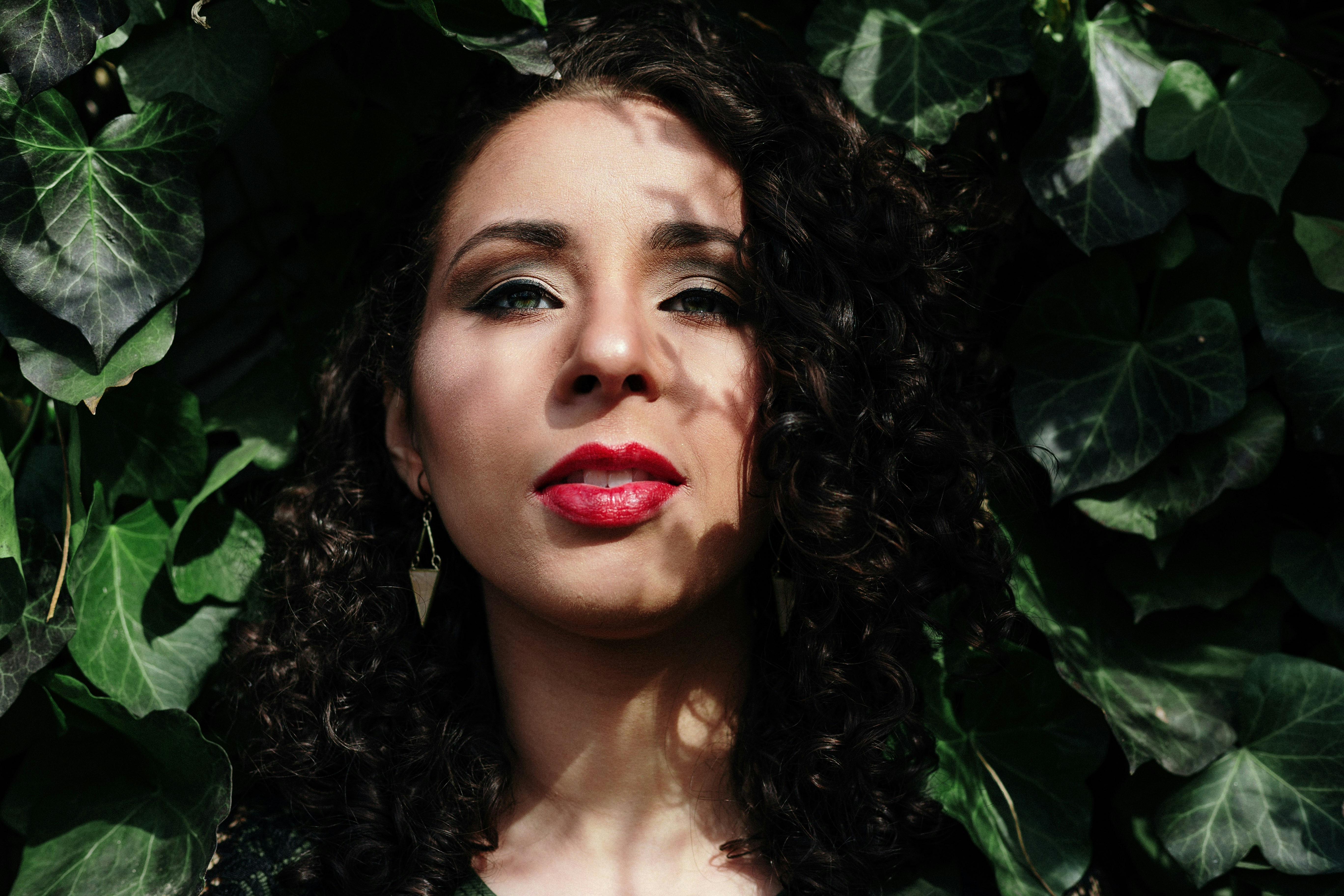 Woman with curly hair and bold red lips stands against a backdrop of lush green leaves, exuding confidence and connection to nature.