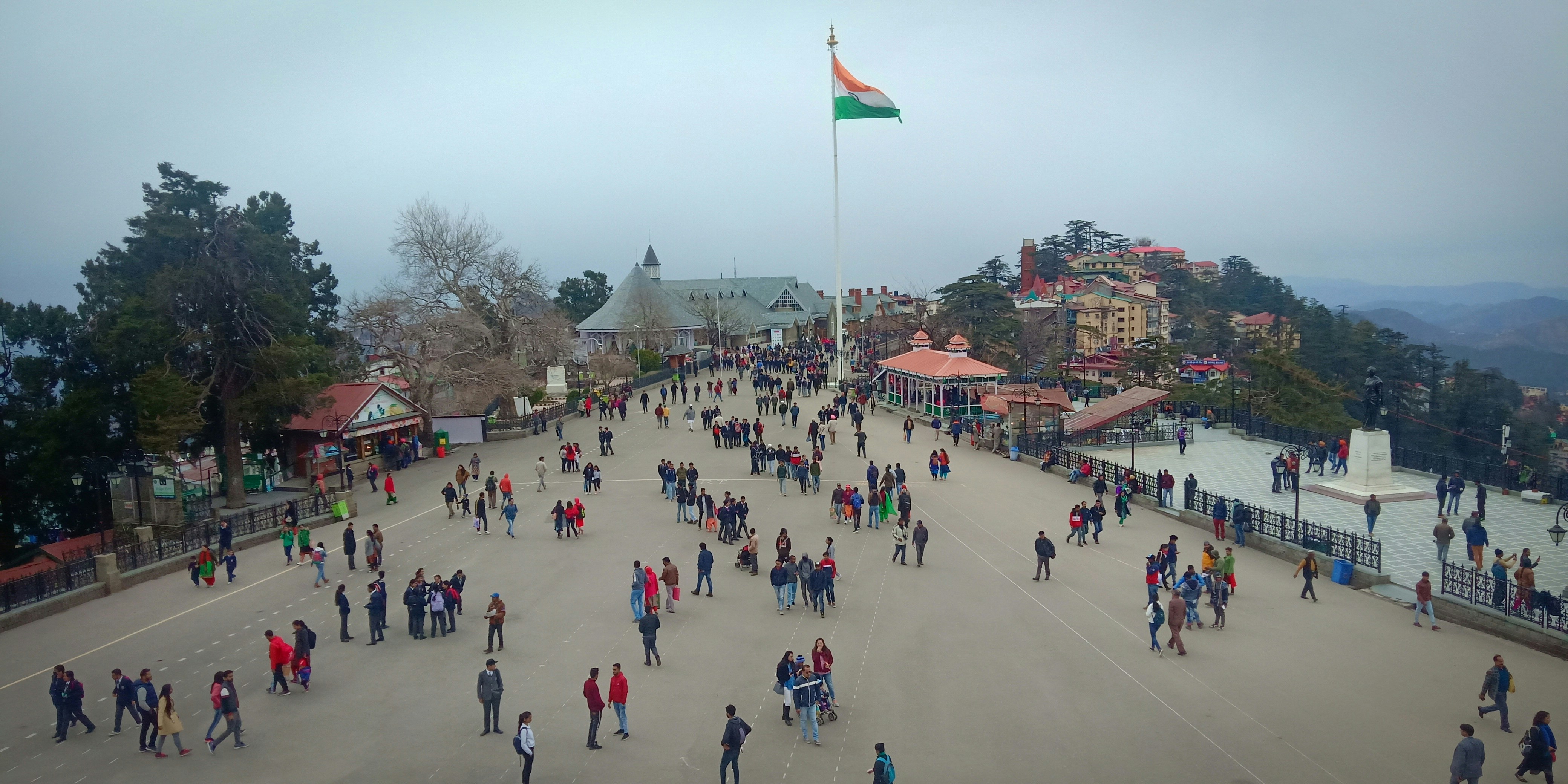 Crowd of visitors enjoying a bustling promenade beneath a large Indian flag, surrounded by scenic hills and architecture.
