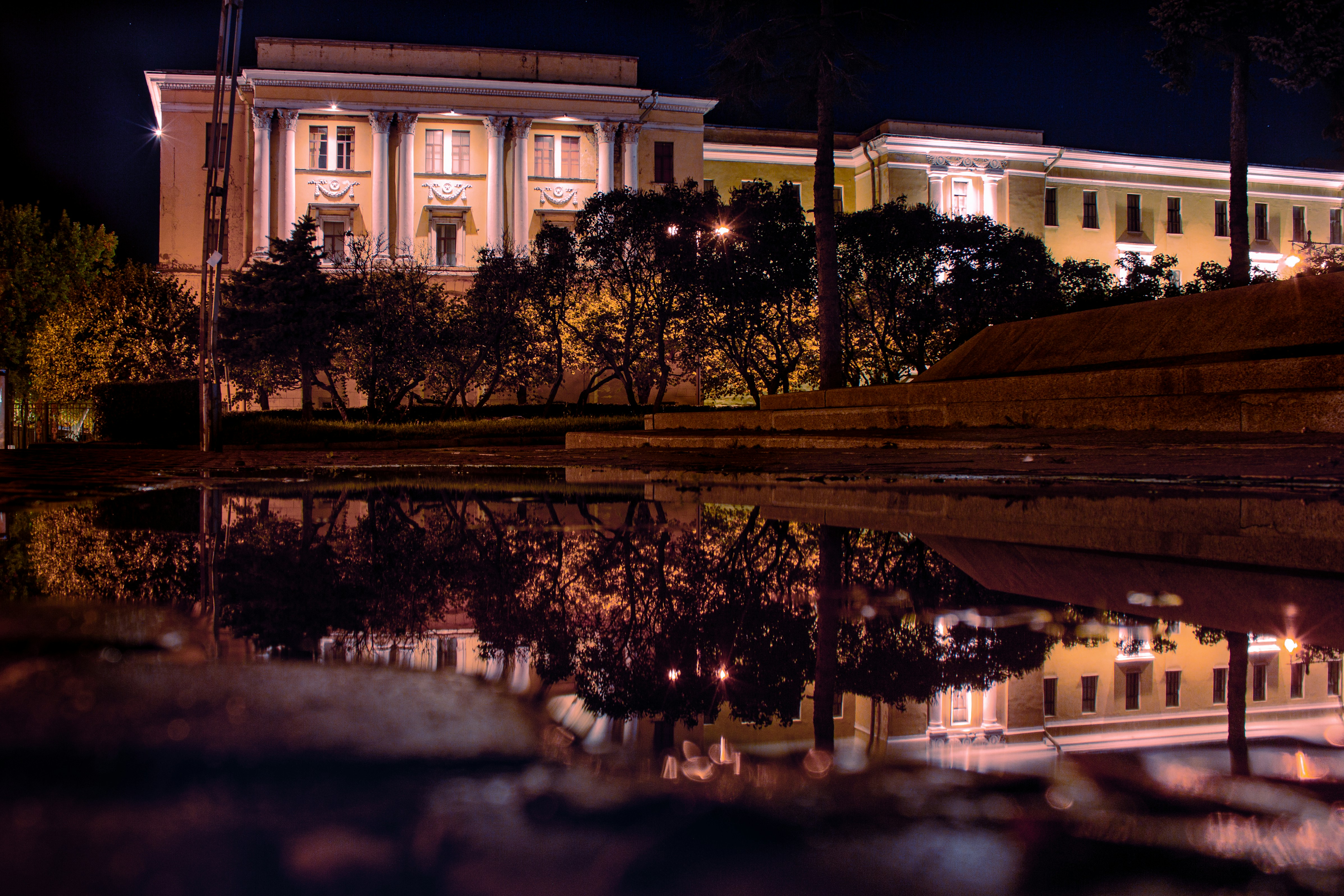 Historic building illuminated at night, reflected in a puddle on cobblestone pavement.