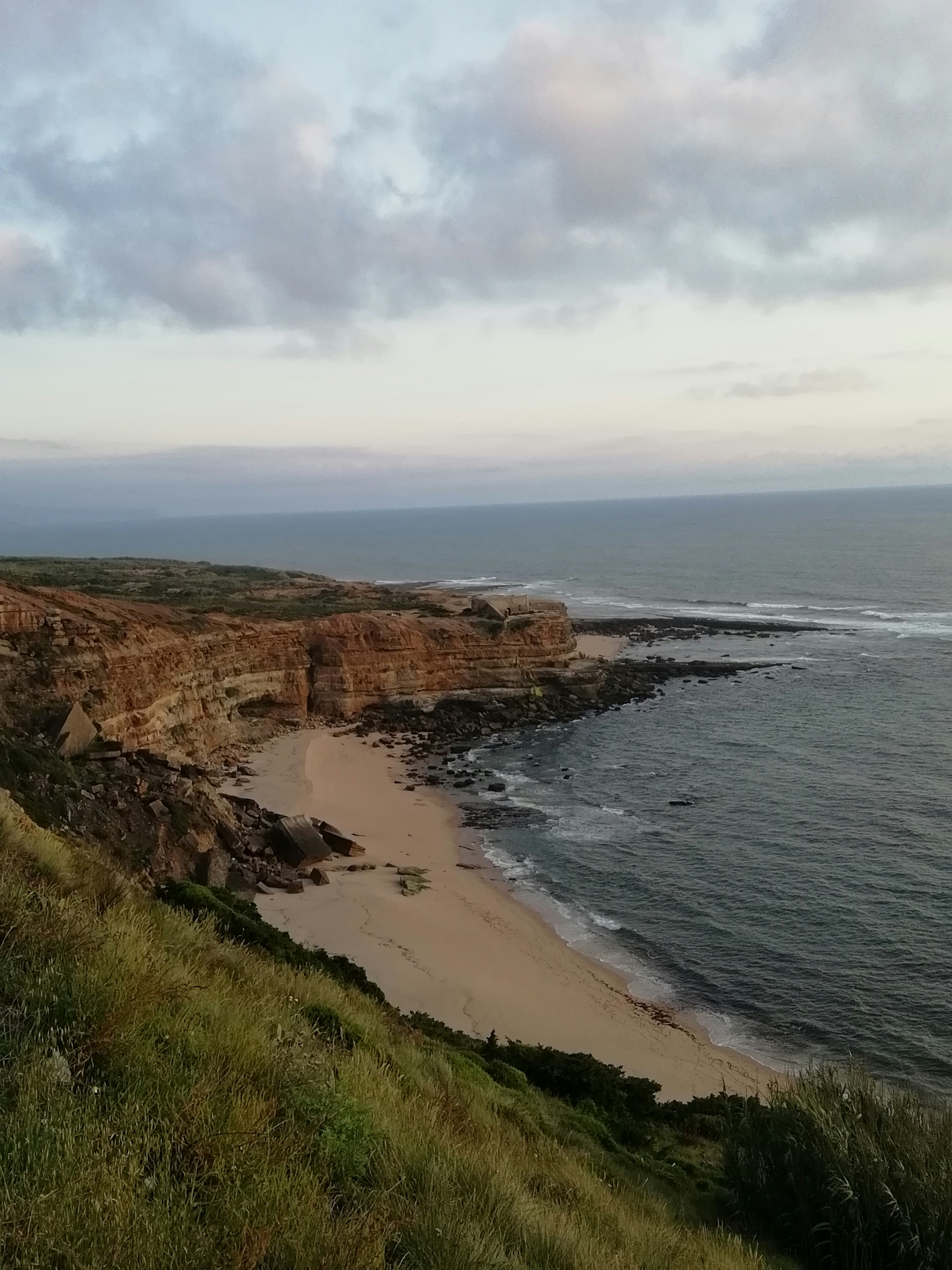 Coastal cliffs frame a sandy cove and rocky shoreline while the calm sea stretches to the horizon. A landscape photograph capturing rugged geology and maritime quiet.