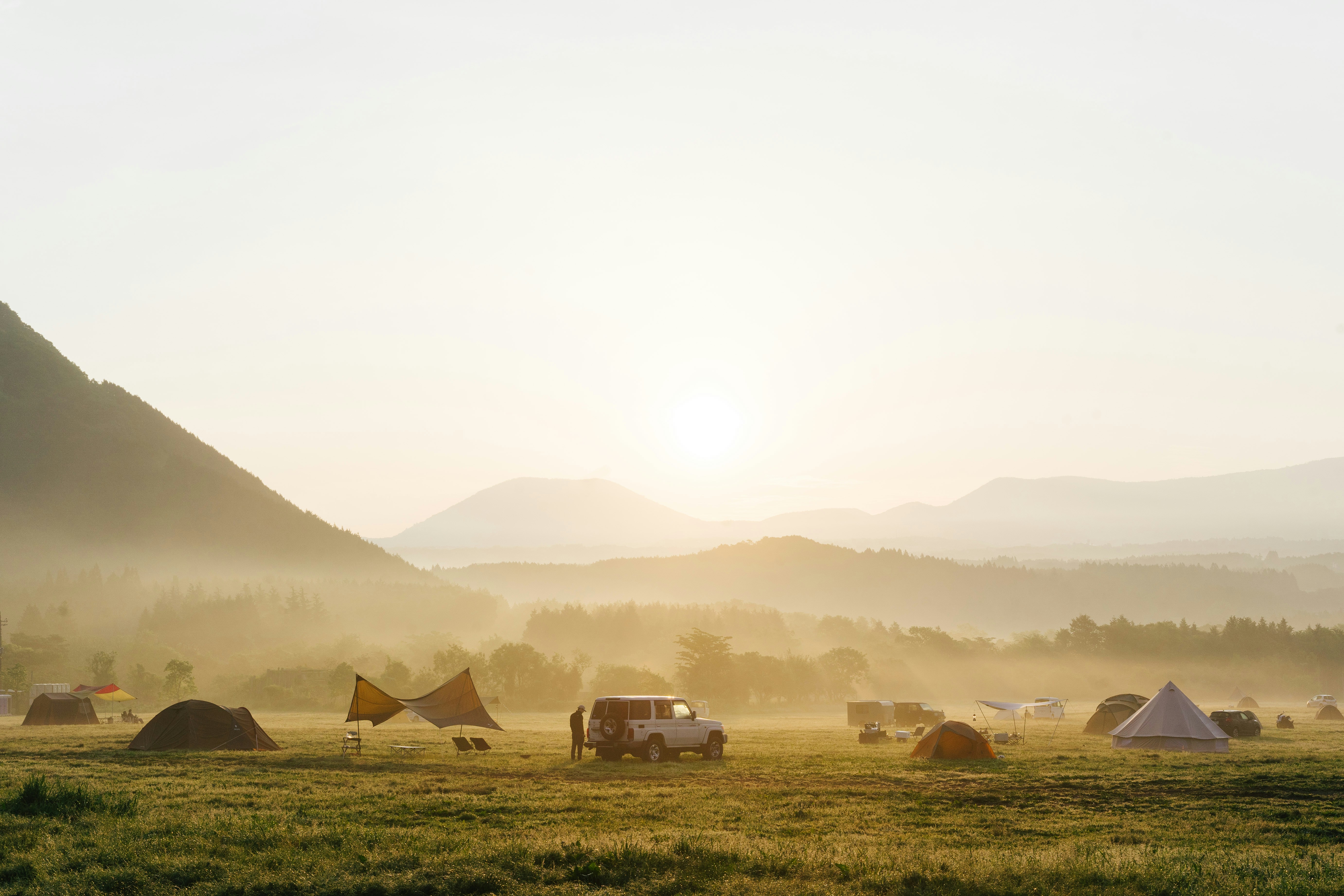Tents and a vehicle on a grassy field with mountains in the background at sunrise.