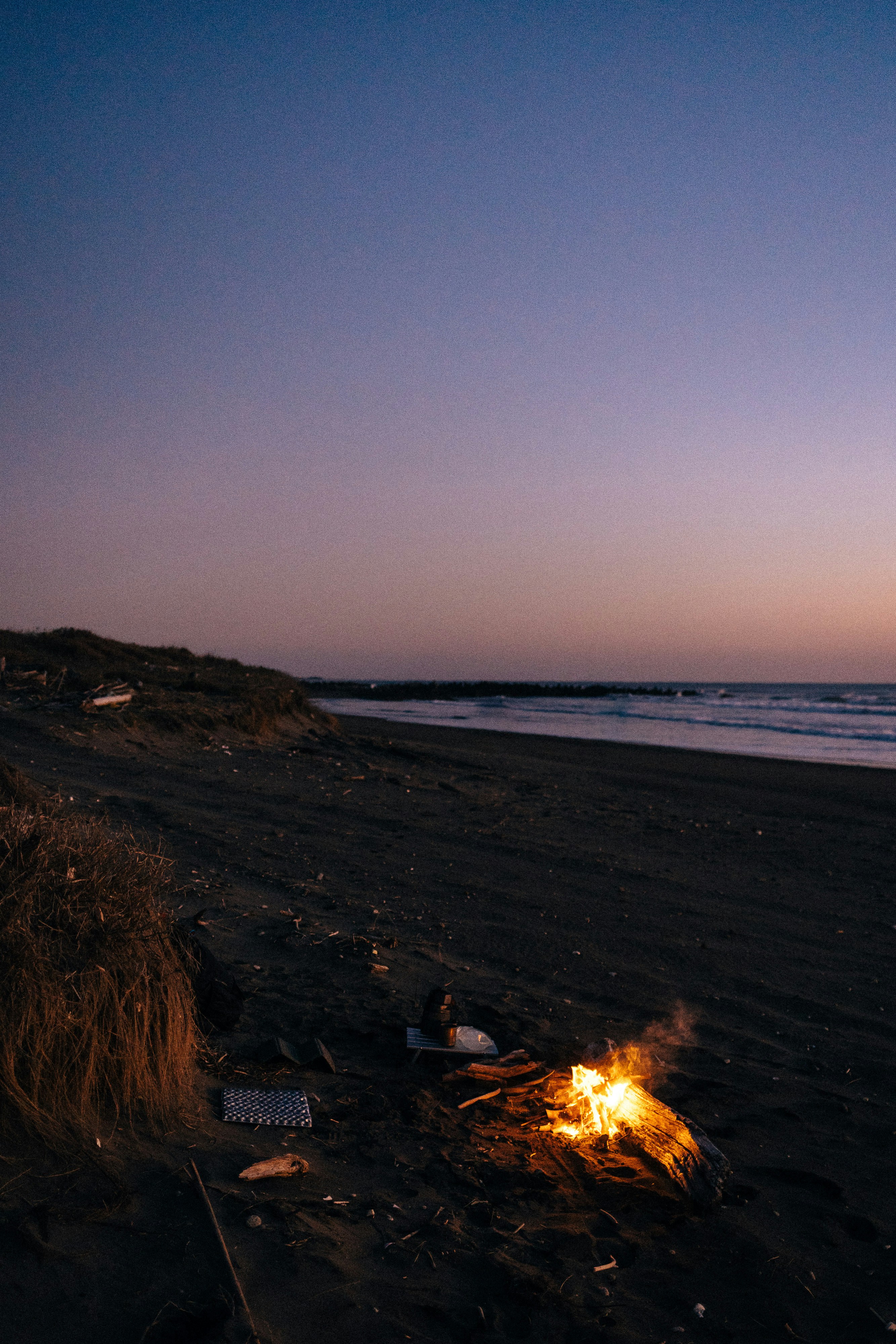 A small campfire flickers on the beach, surrounded by scattered driftwood, as twilight descends over the calm ocean waves.