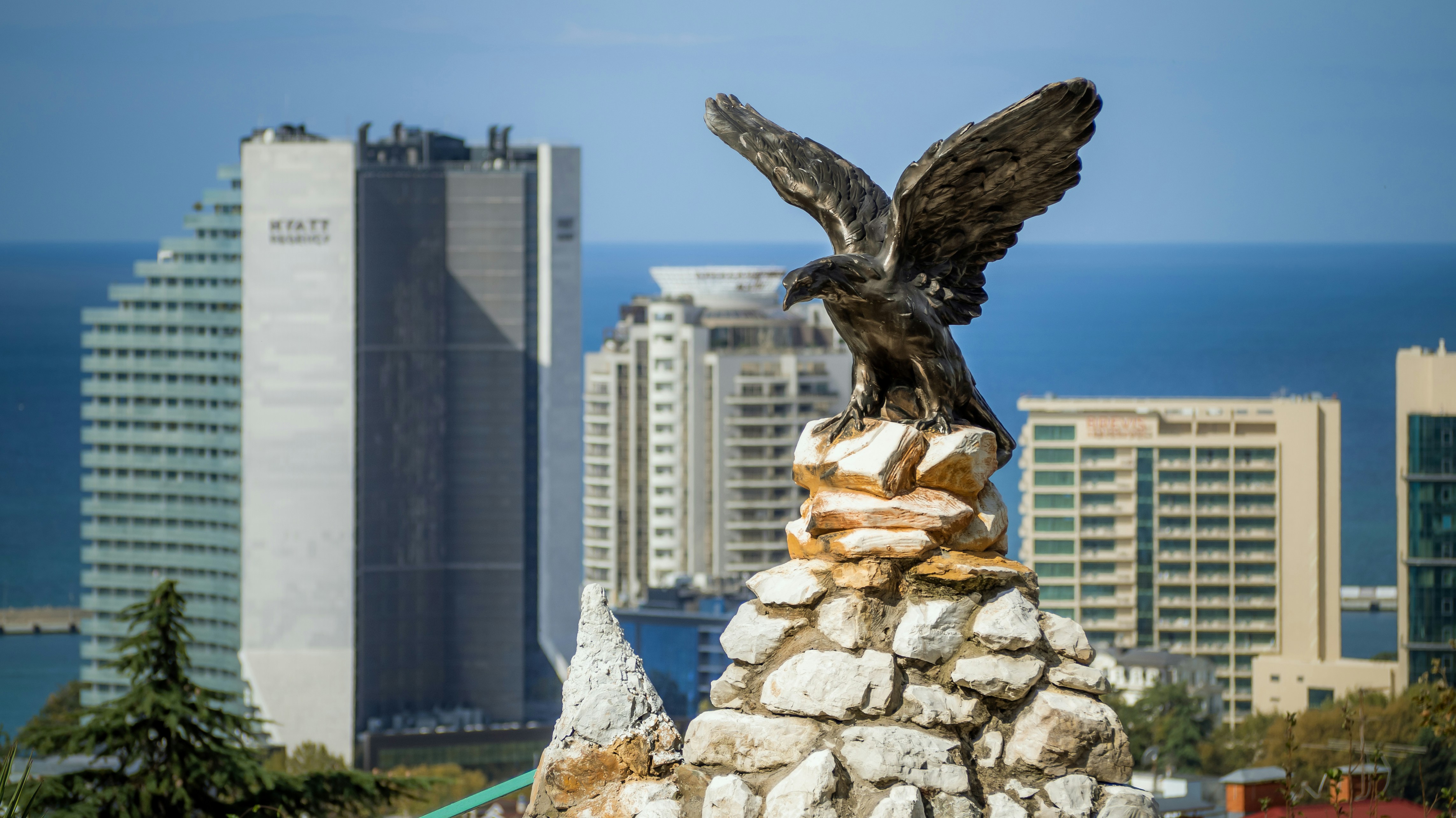 Bronze eagle statue perched atop a stone base, overlooking a modern city skyline by the sea.