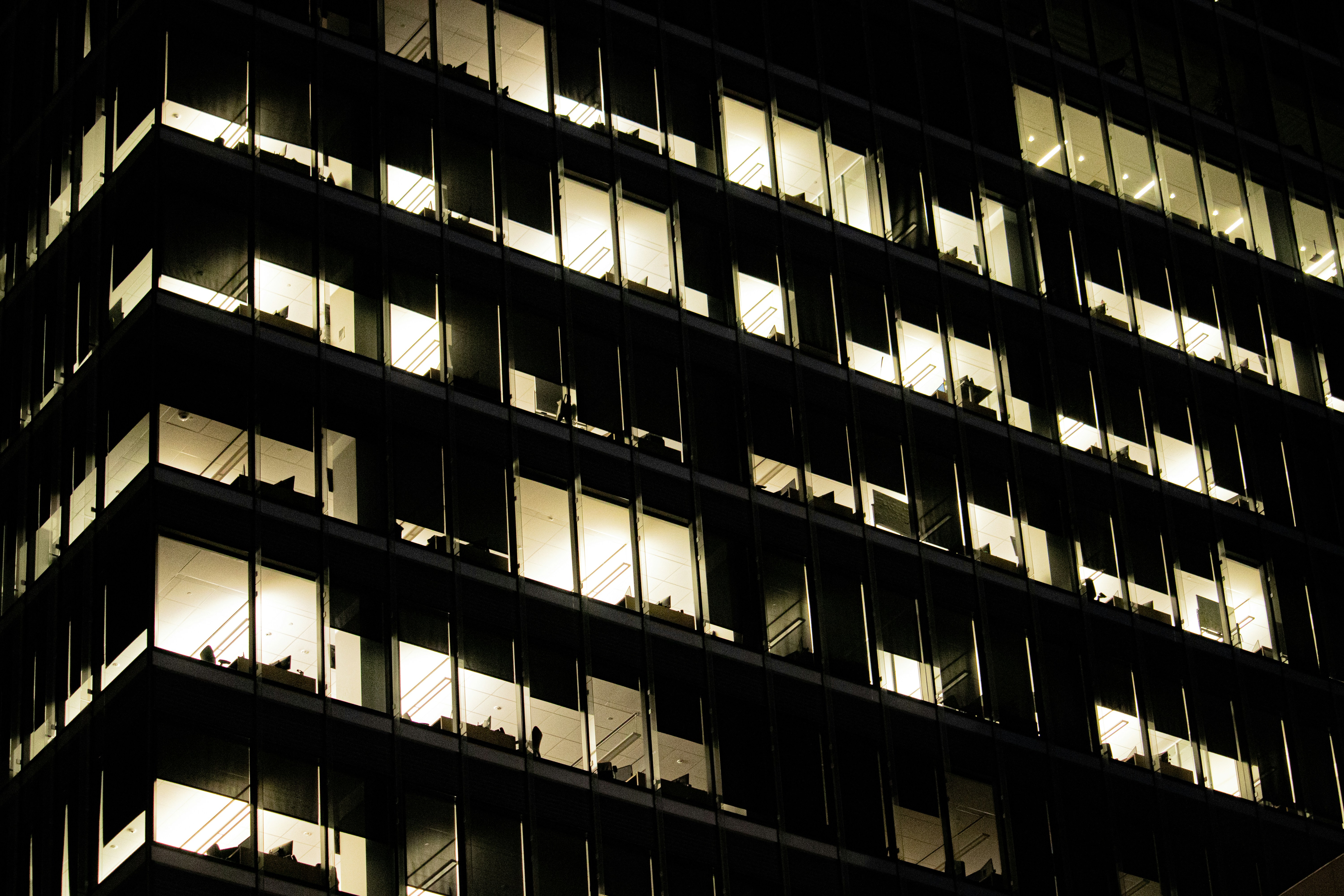 Illuminated office windows reveal silhouettes of workers in a modern skyscraper at night.