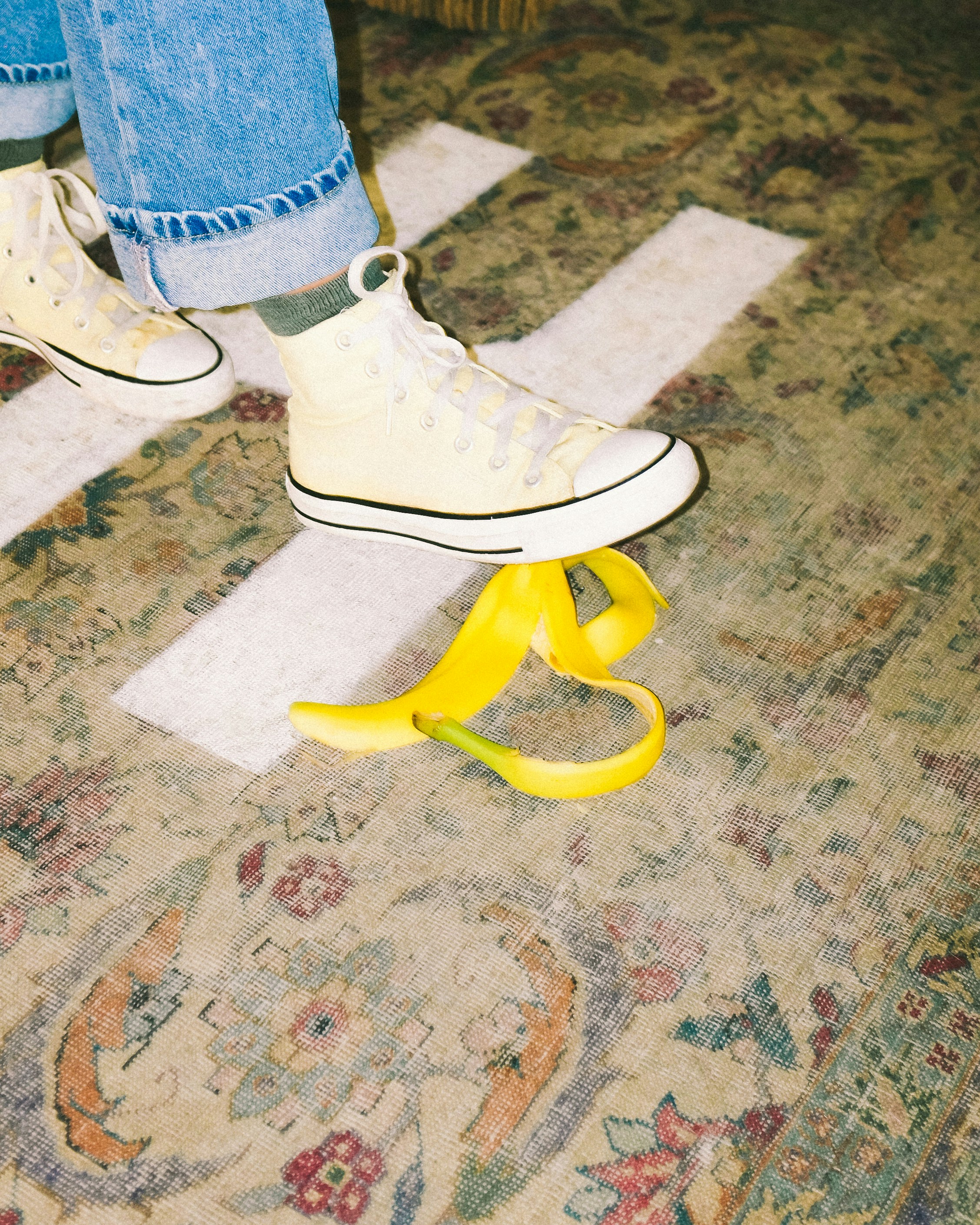 A pair of yellow sneakers precariously positioned on a banana peel, set against a patterned rug and painted white stripes.