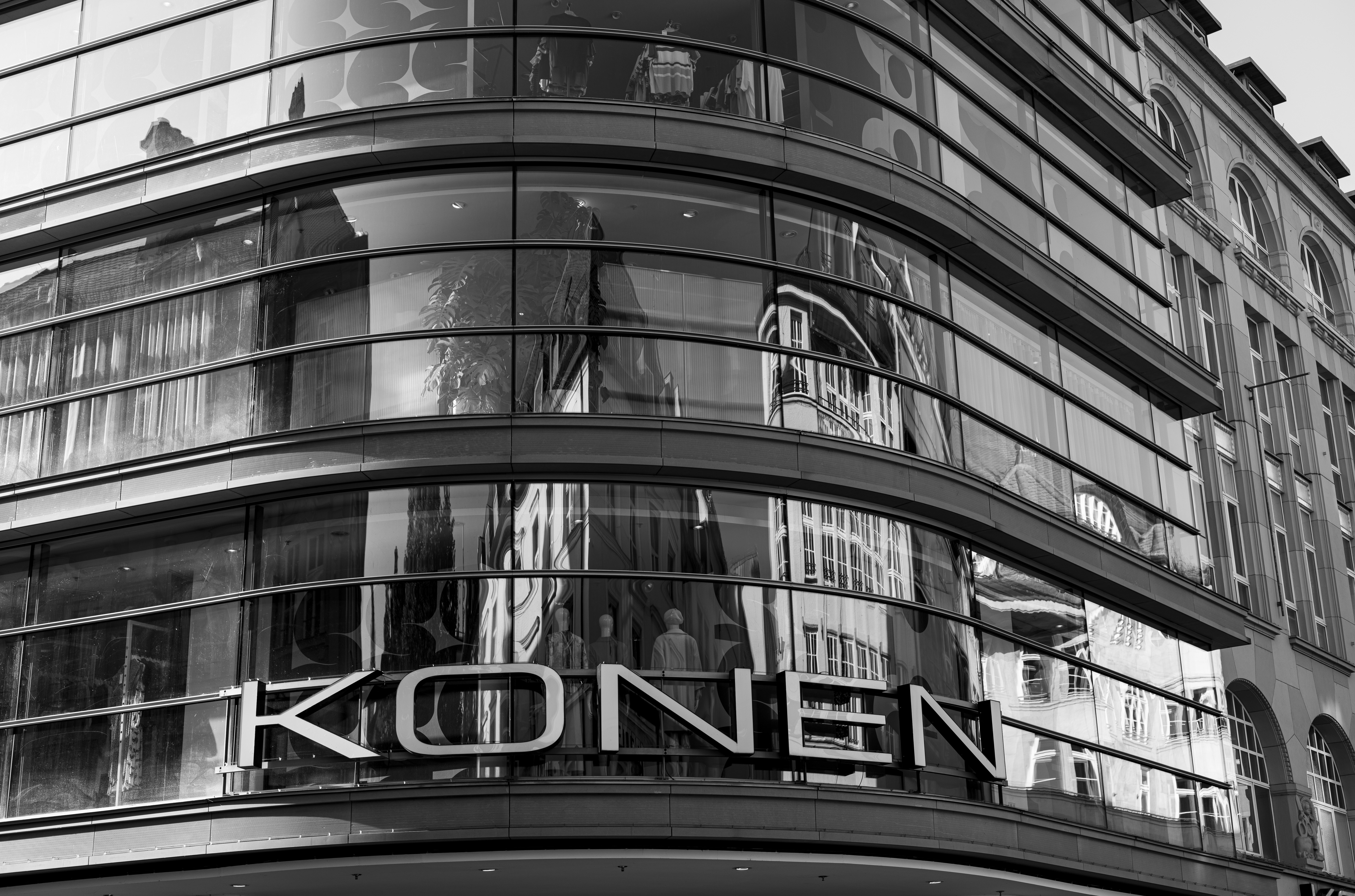 Modern architectural facade featuring the name 'KONEN' with reflections of surrounding buildings in glass. The monochrome palette adds a timeless quality.