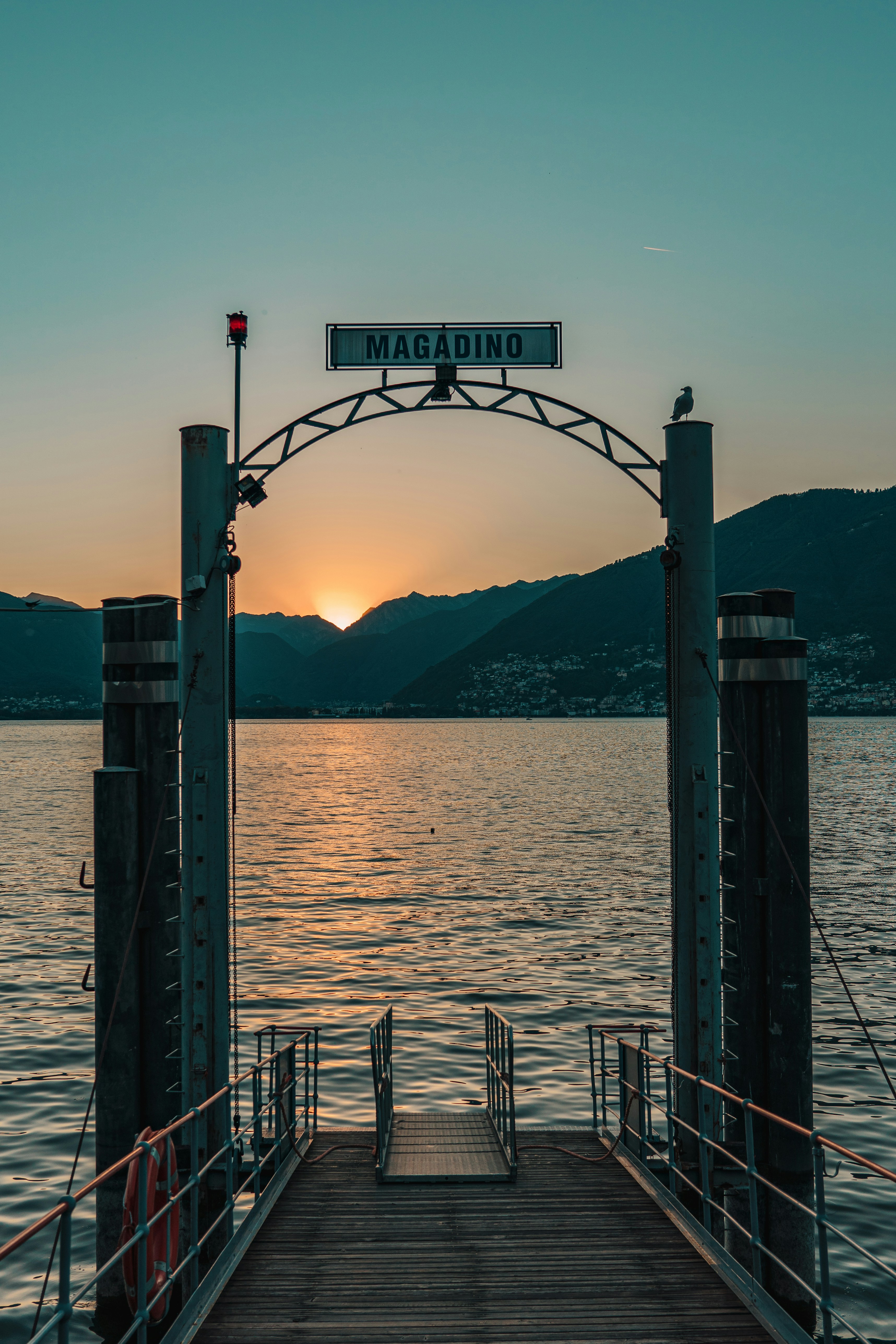 Pier at Magadino silhouetted against a vibrant sunset, with mountains framing the horizon.