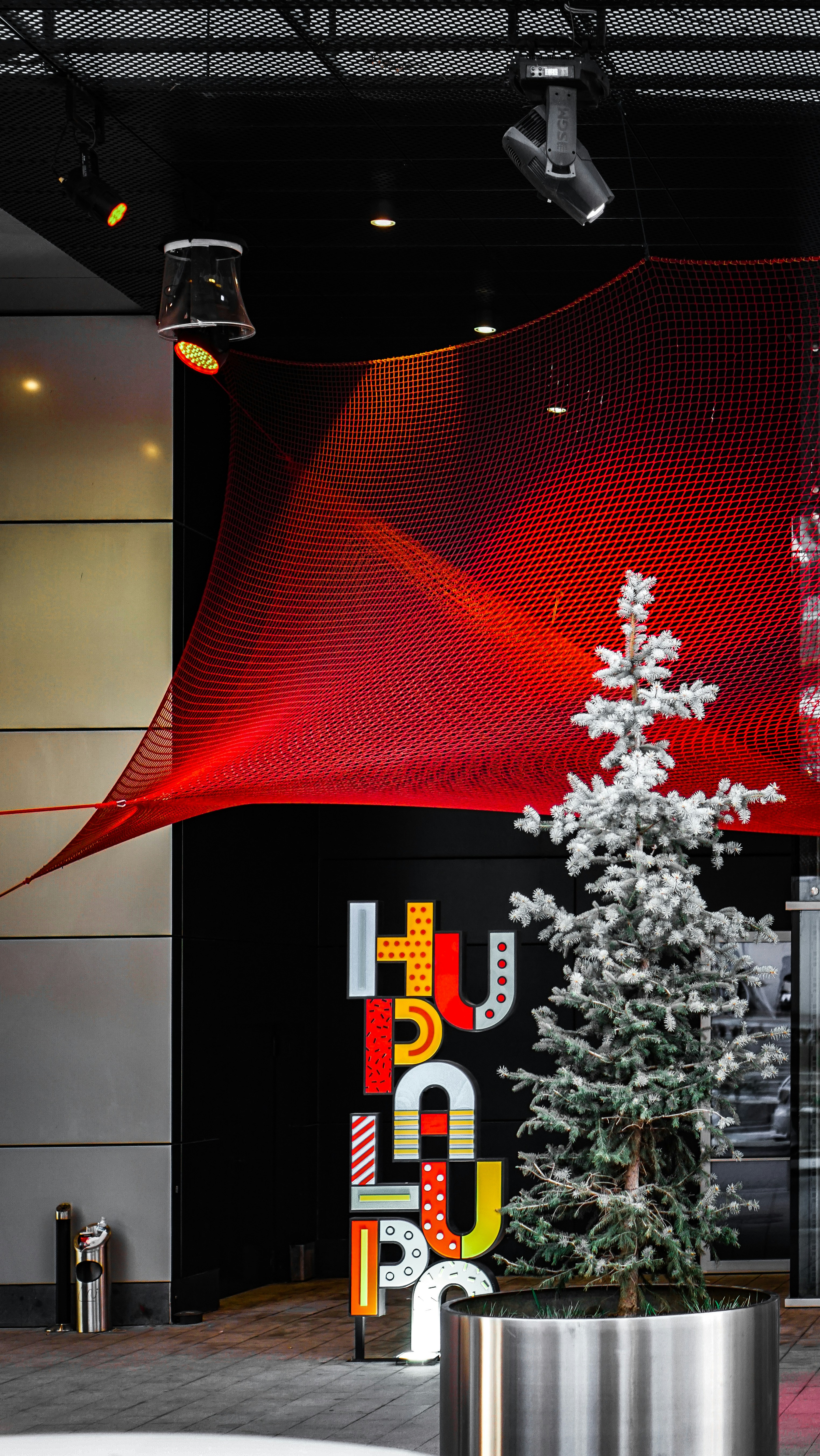 Crimson Mesh Over a Colorful Letter Sculpture Beside a Snow-Dusted Indoor Tree