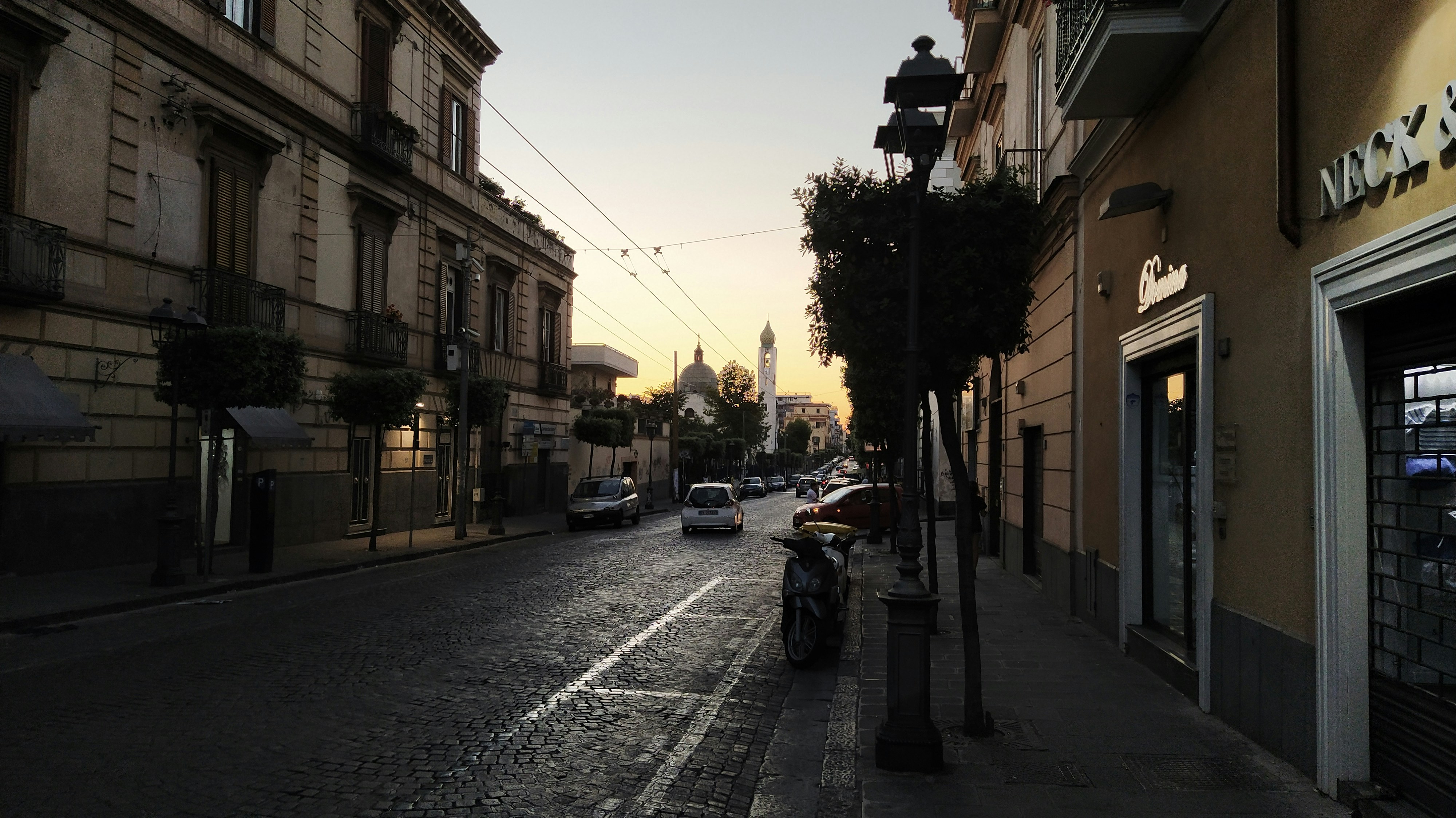 City street lined with parked cars and historic buildings under a twilight sky.