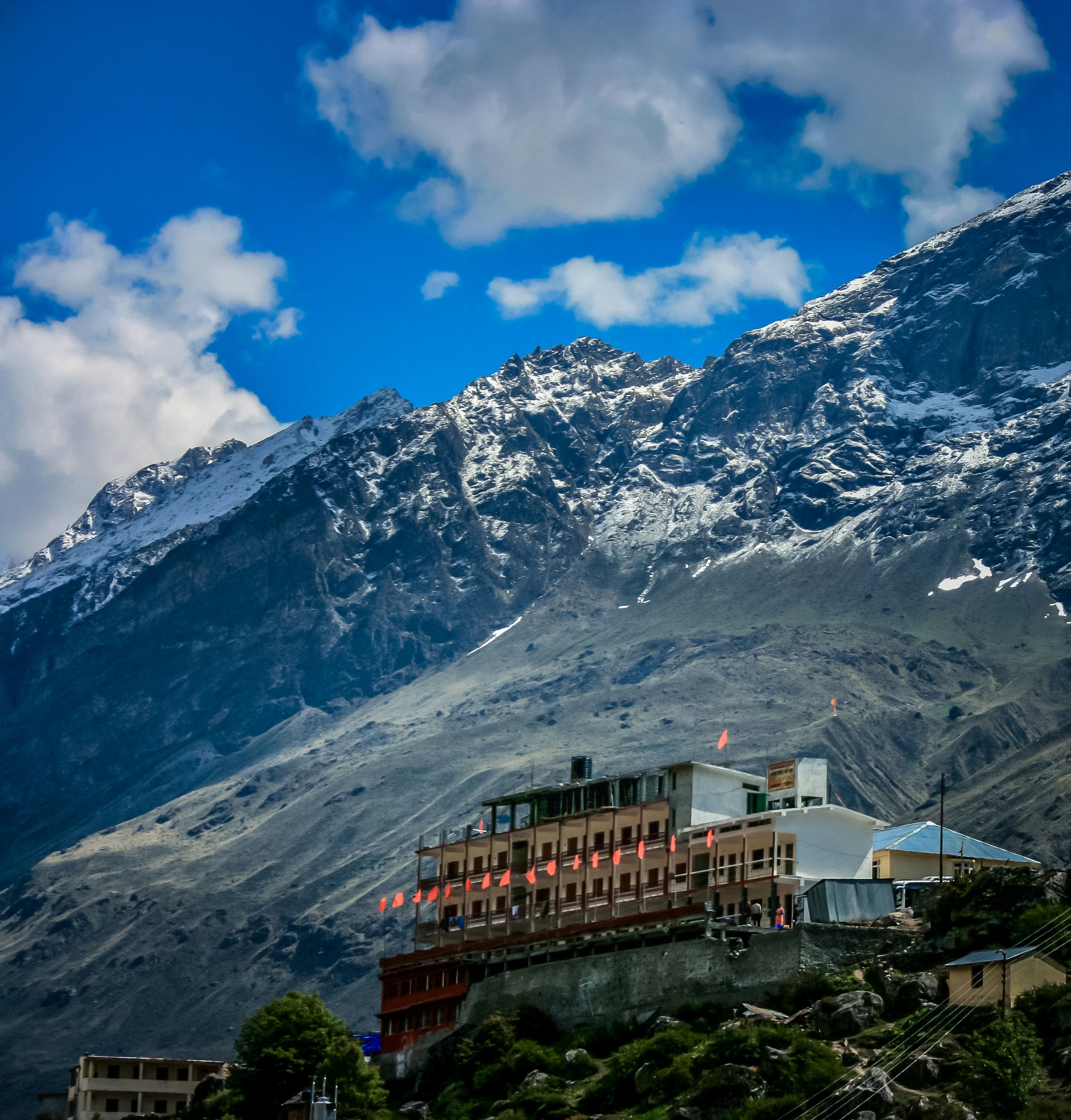 A mountain lodge perched on a rocky hillside, surrounded by towering snow-capped peaks and a clear blue sky with fluffy clouds.