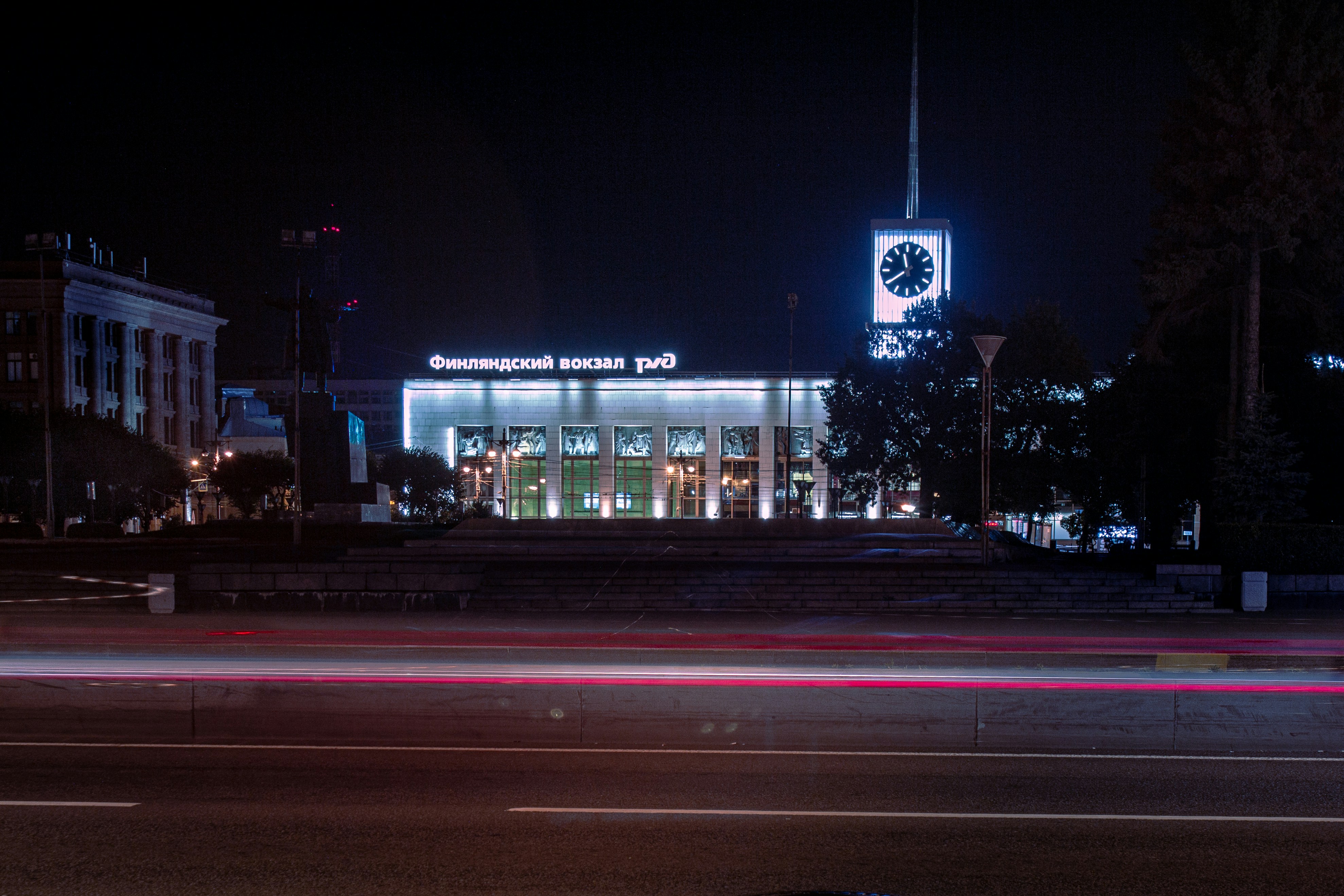 Illuminated railway station facade featuring modern architecture against a dark sky, with light trails from passing vehicles. 