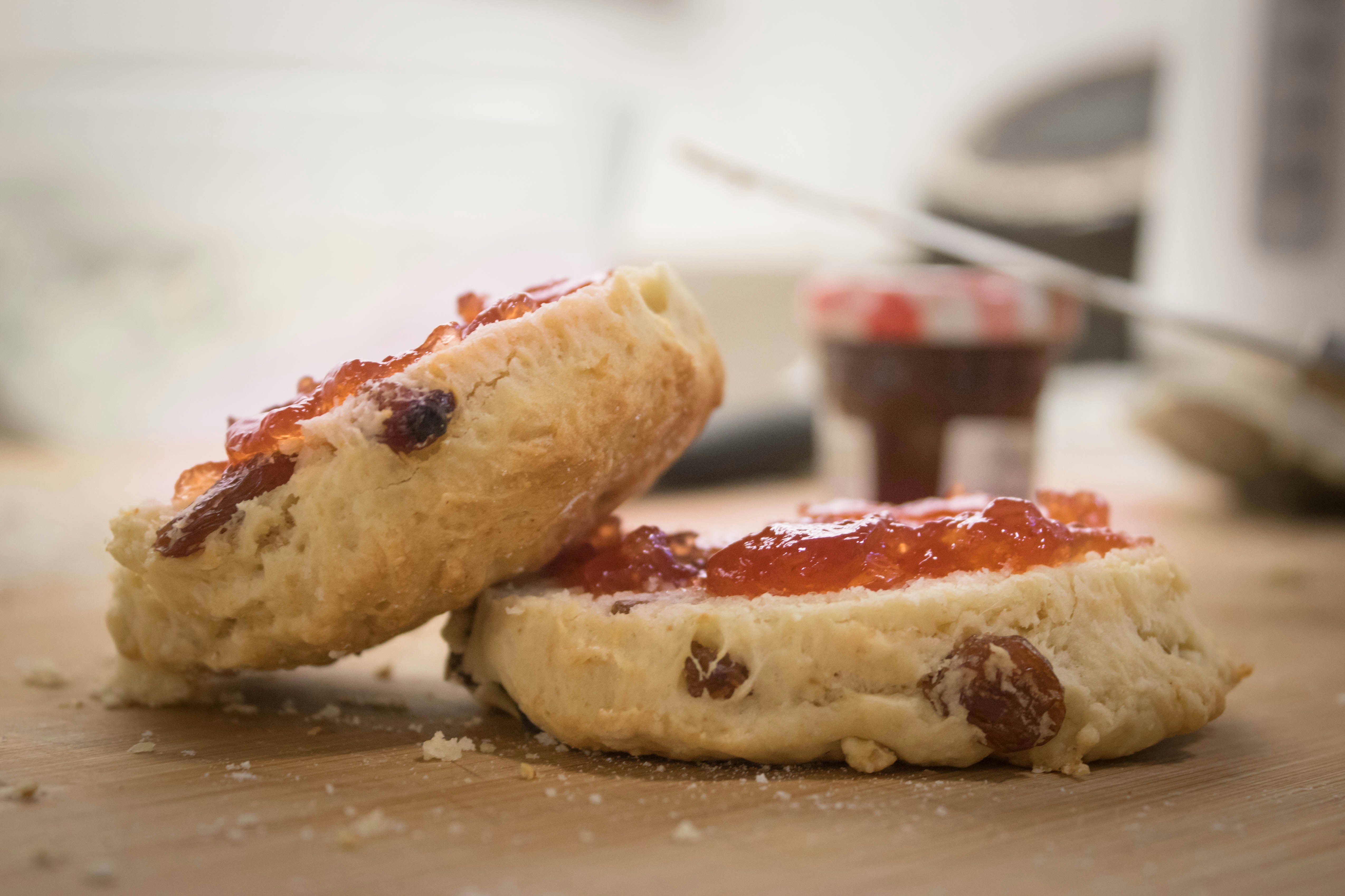 Two freshly baked scones topped with vibrant strawberry jam, resting on a wooden surface with a jar of jam in the background.