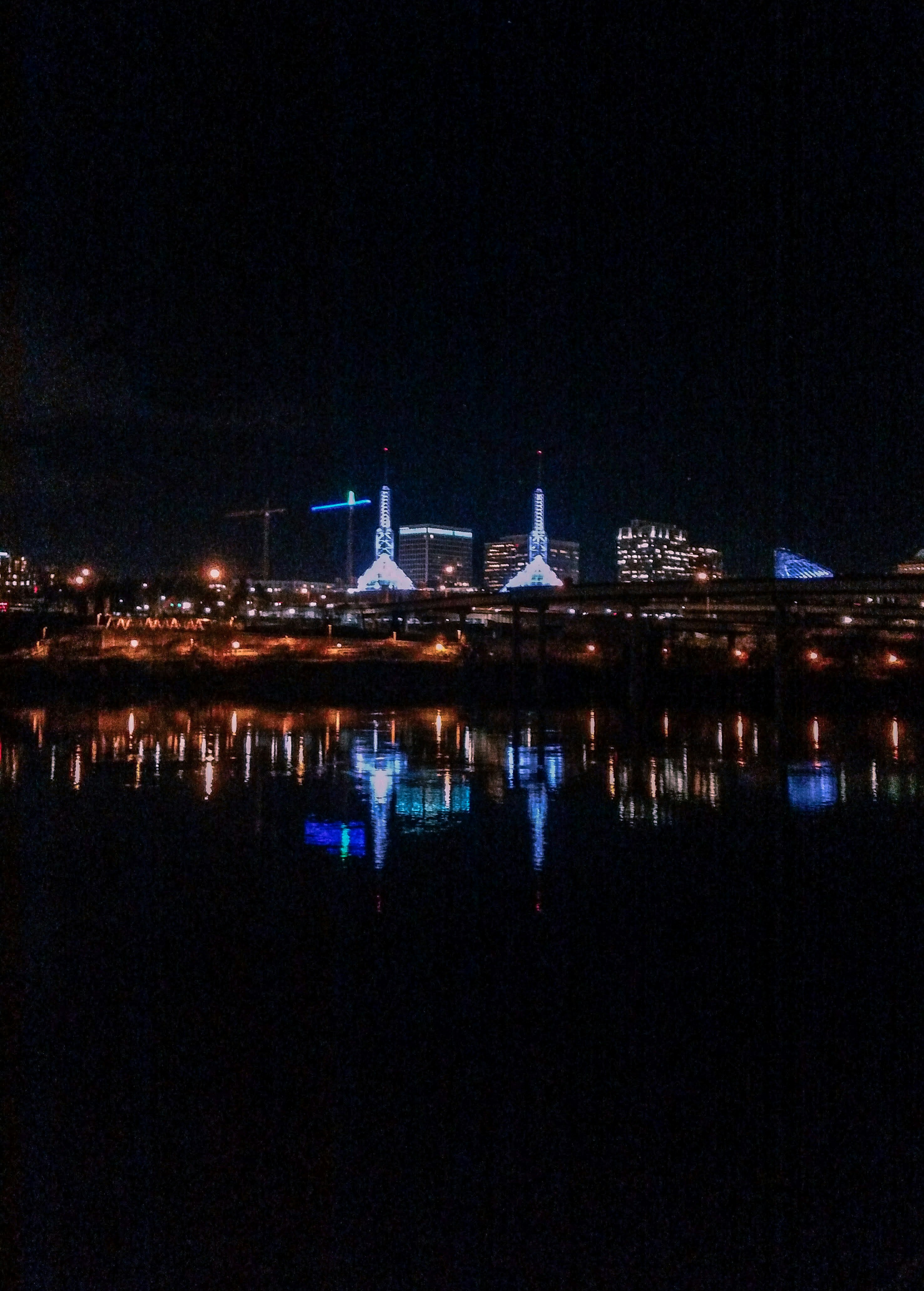 Night city skyline over a dark river with blue-lit towers and their reflections on the water. A city-night photograph.
