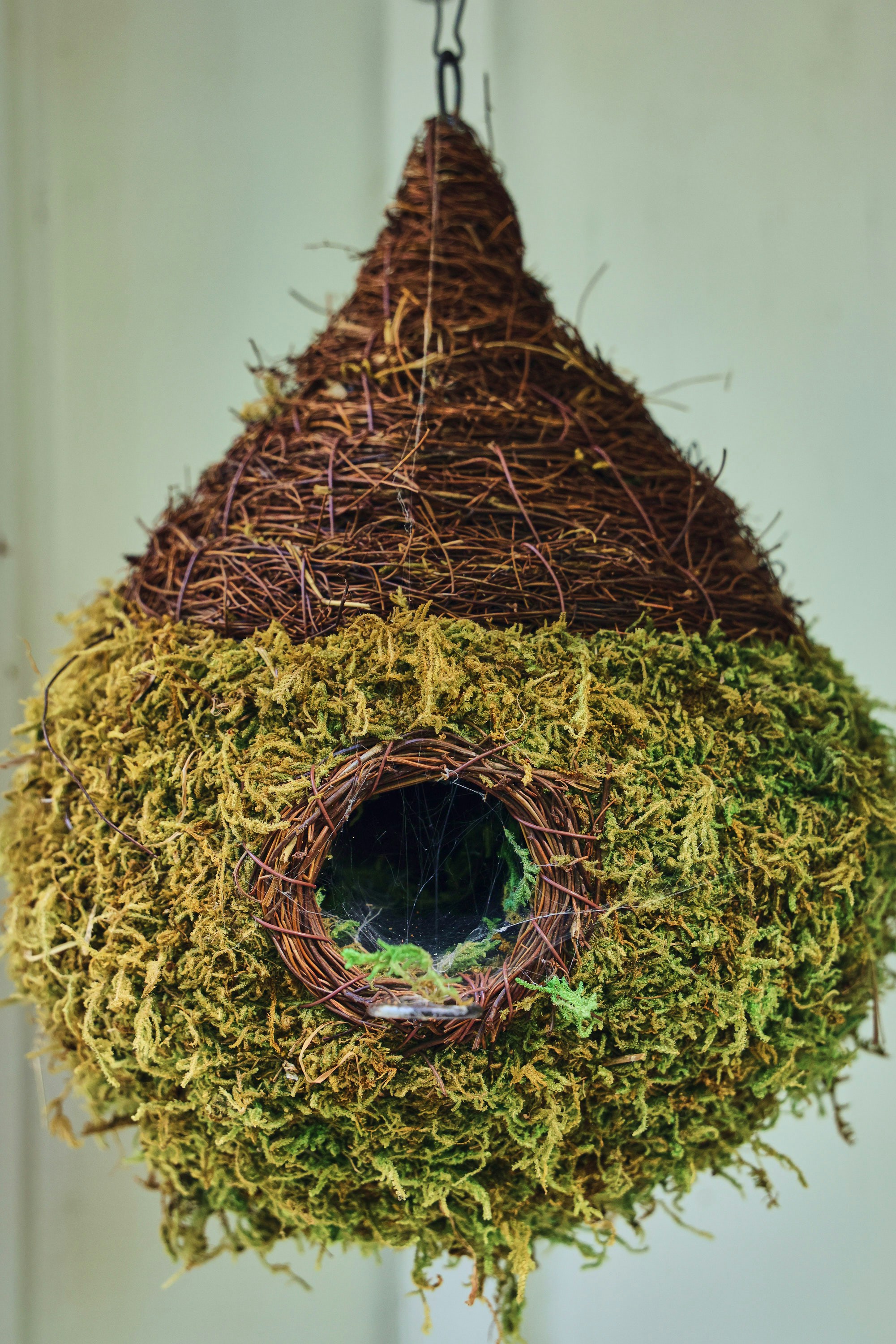 Intricately woven birdhouse adorned with moss and twigs, featuring a circular entrance, suspended against a soft backdrop.