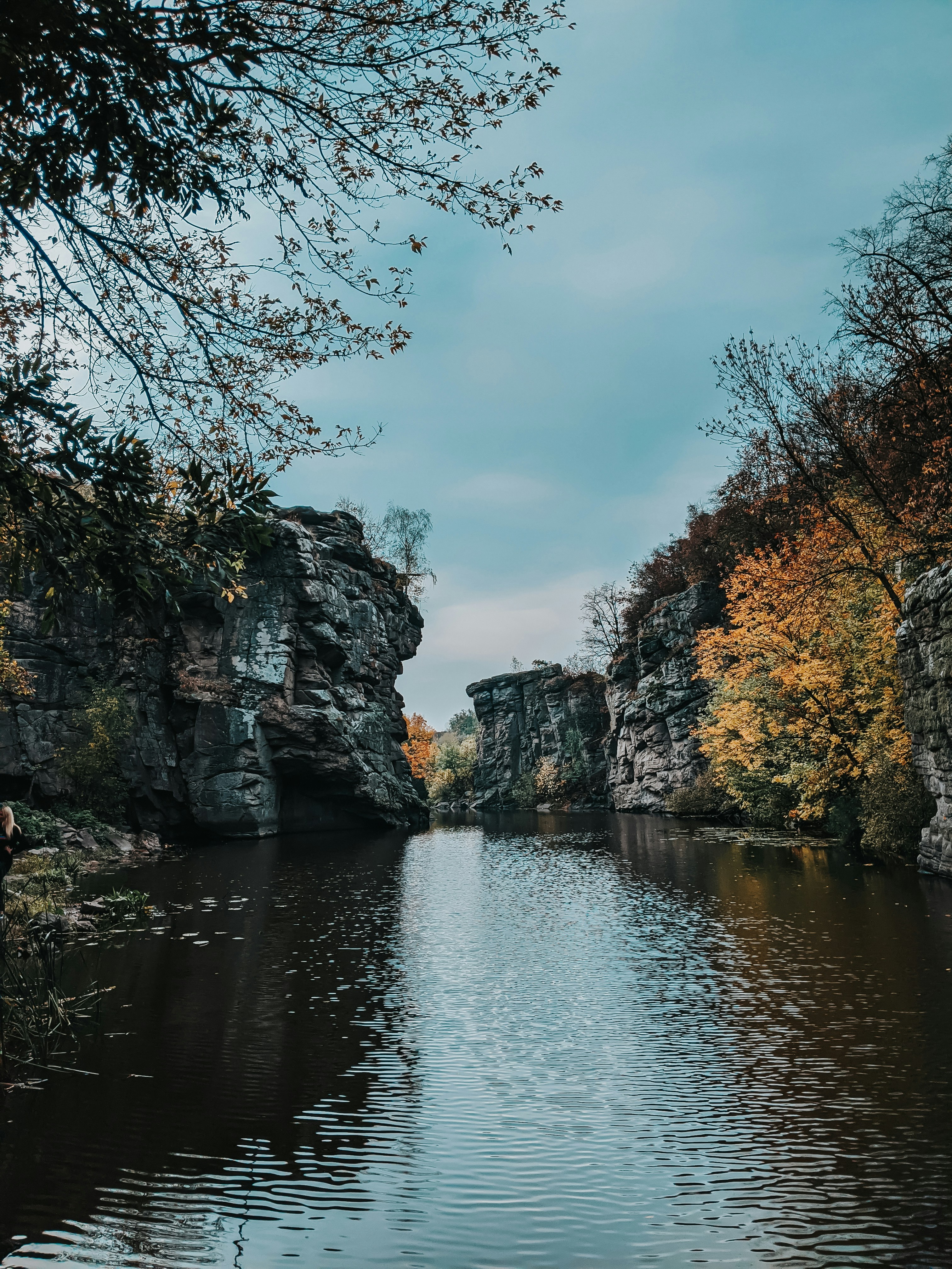 A tranquil river flows between towering rock formations adorned with vibrant autumn foliage. The scene captures the peaceful coexistence of nature's elements.