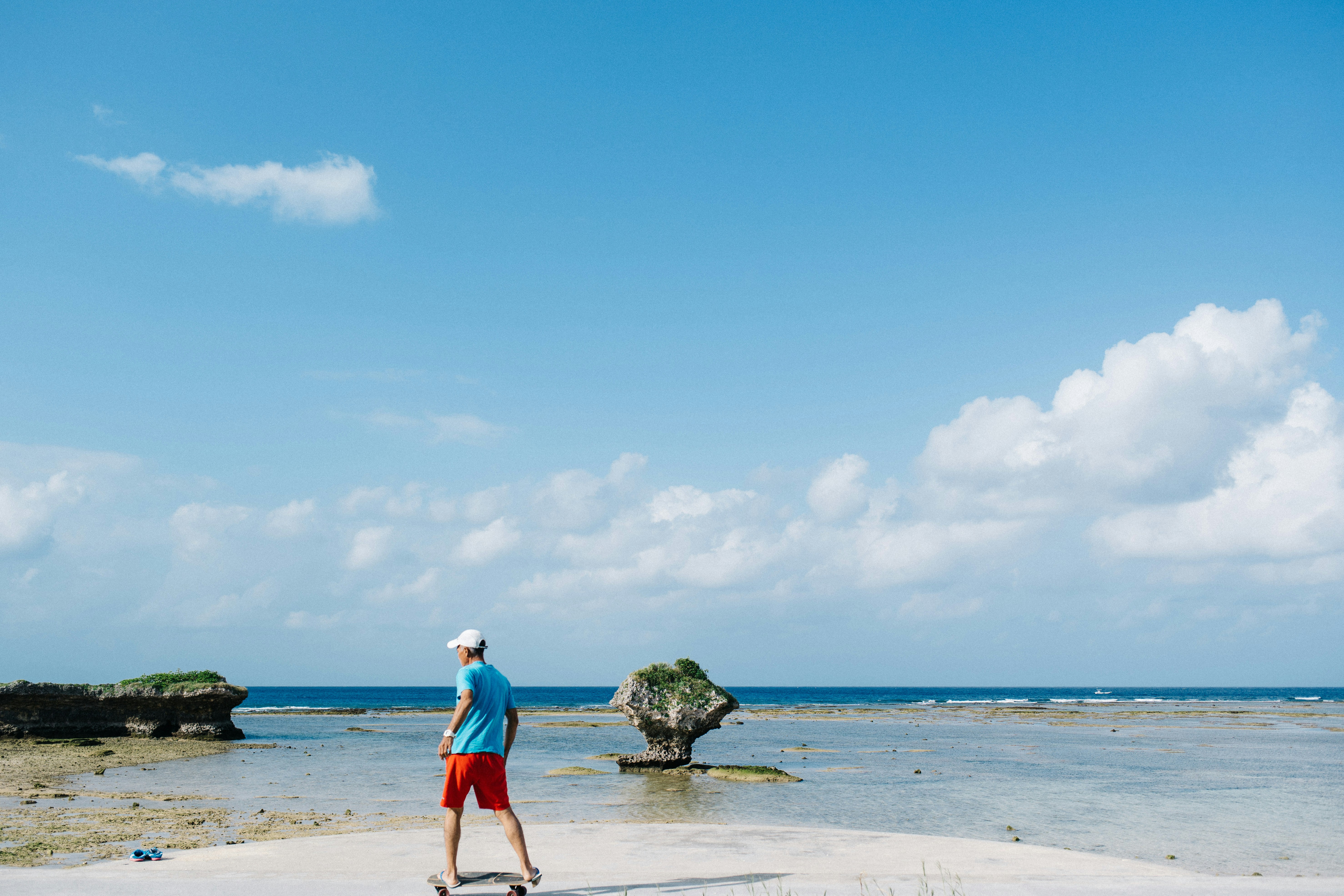 A lone figure strolls along a coastal pathway, with a unique rock formation rising from the ocean in the background under a bright blue sky.