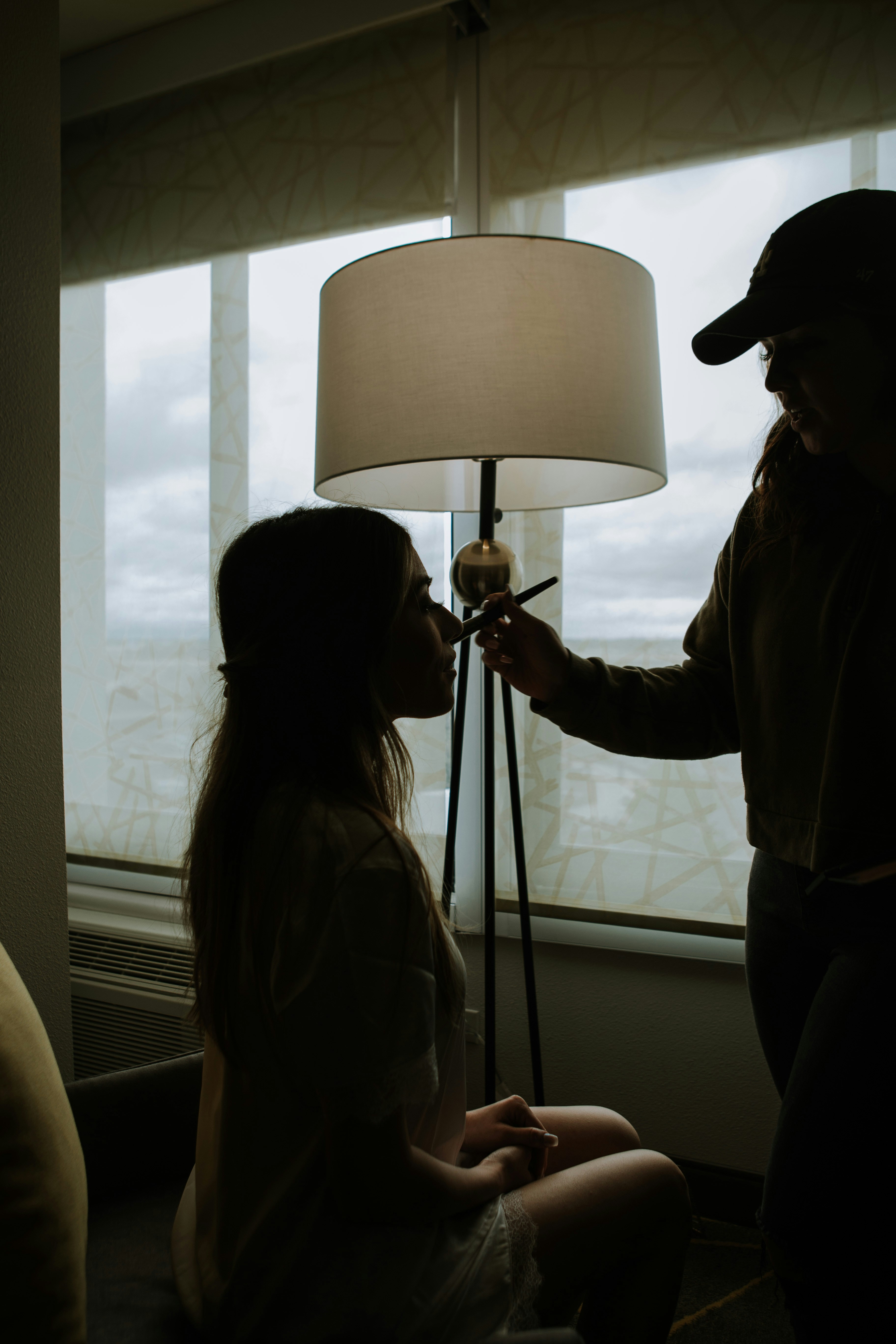 Candid photograph of two women by a window; one applies makeup to the other as a lamp glows nearby.